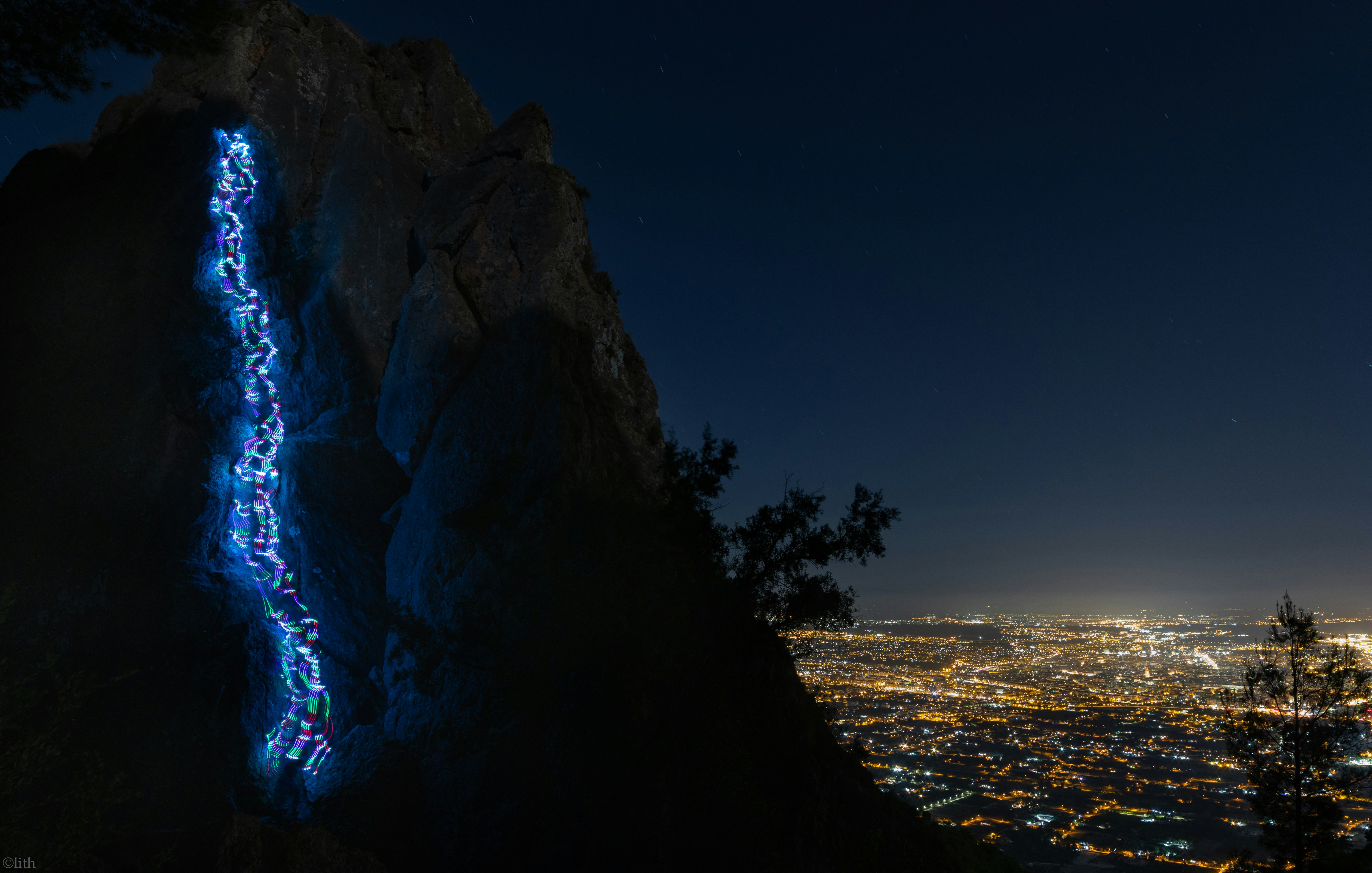 Climber ascends a rocky cliff at night with a cityscape illuminated in the background.