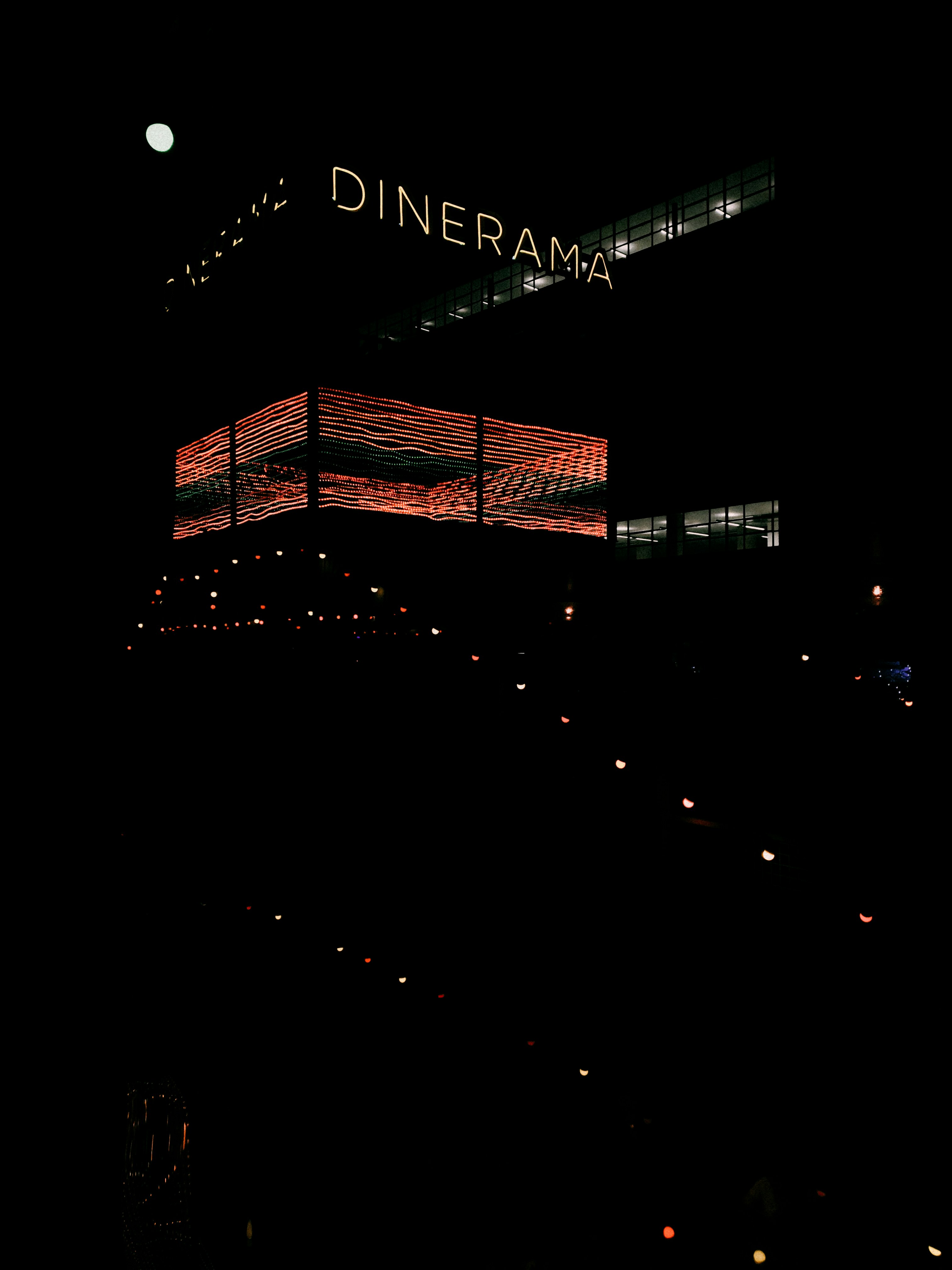 Vibrant neon lights illuminate the exterior of Dinerama, creating a dynamic visual contrast against the night sky. A full moon adds to the ambiance.