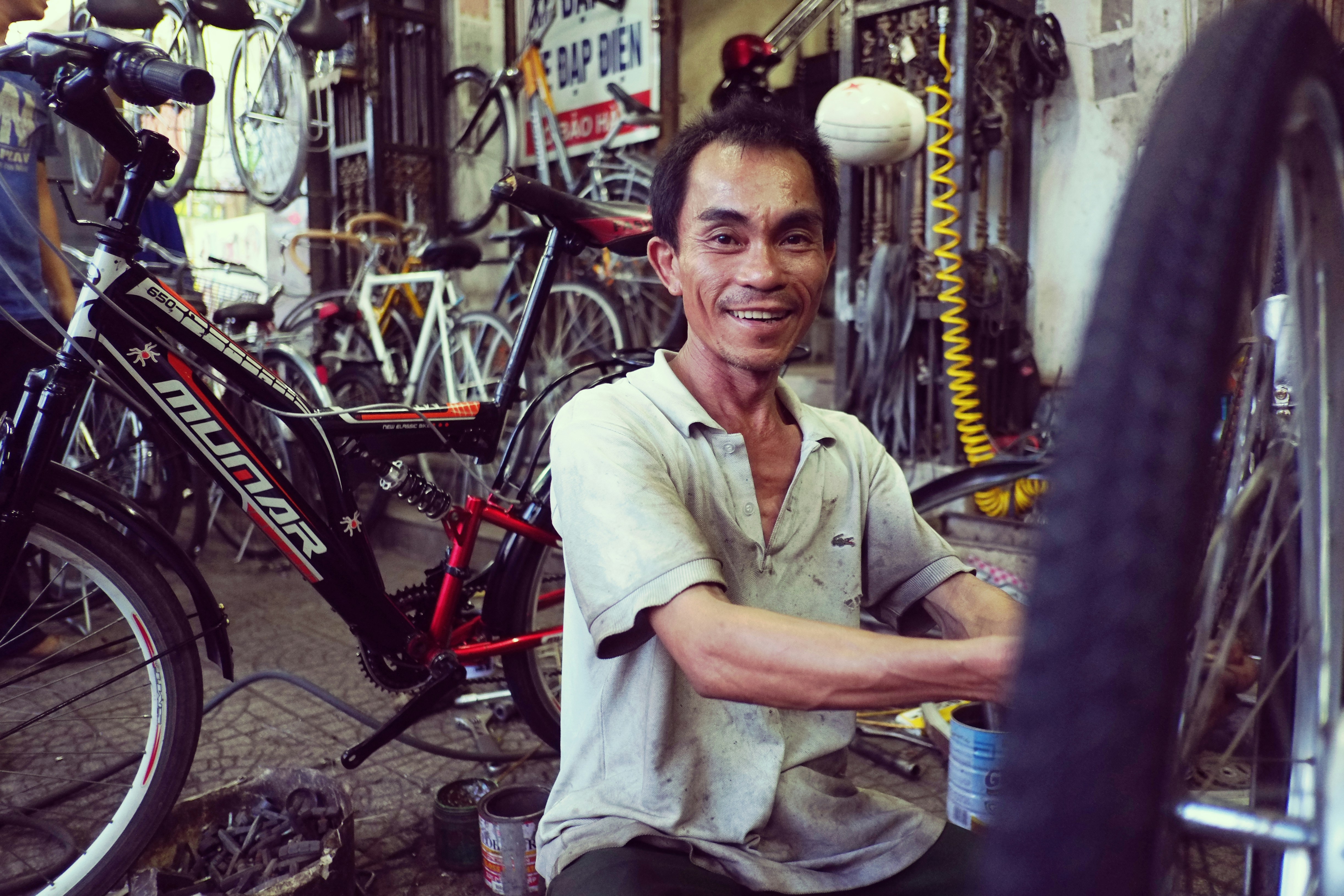 A bicycle mechanic smiles while working on a bike in a cluttered workshop filled with tools and bicycles. The scene captures the essence of craftsmanship and community.