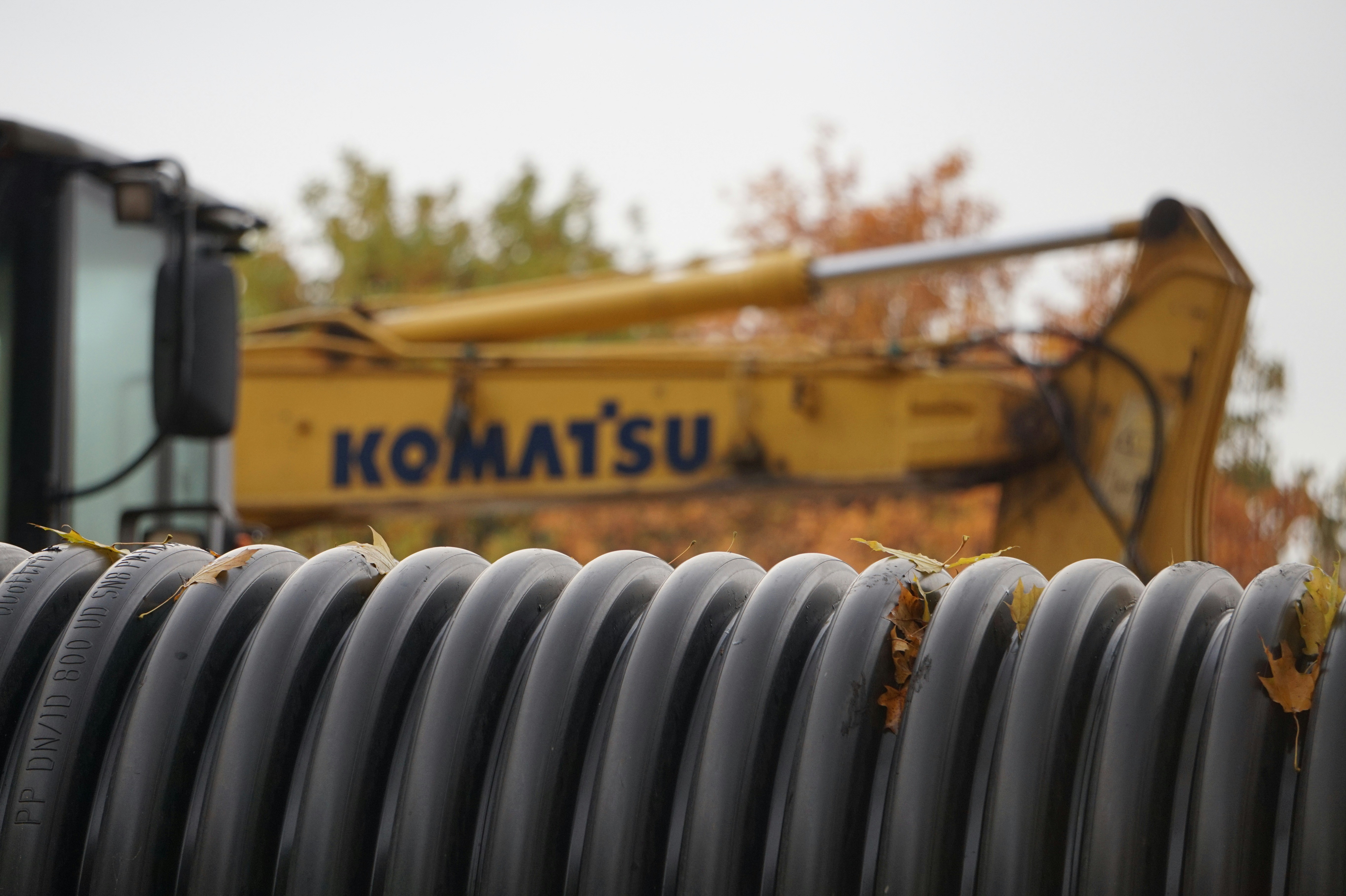 Coiled industrial cables in the foreground with a Komatsu excavator blurred in the background; a scene from a construction site featuring heavy machinery and autumn foliage.