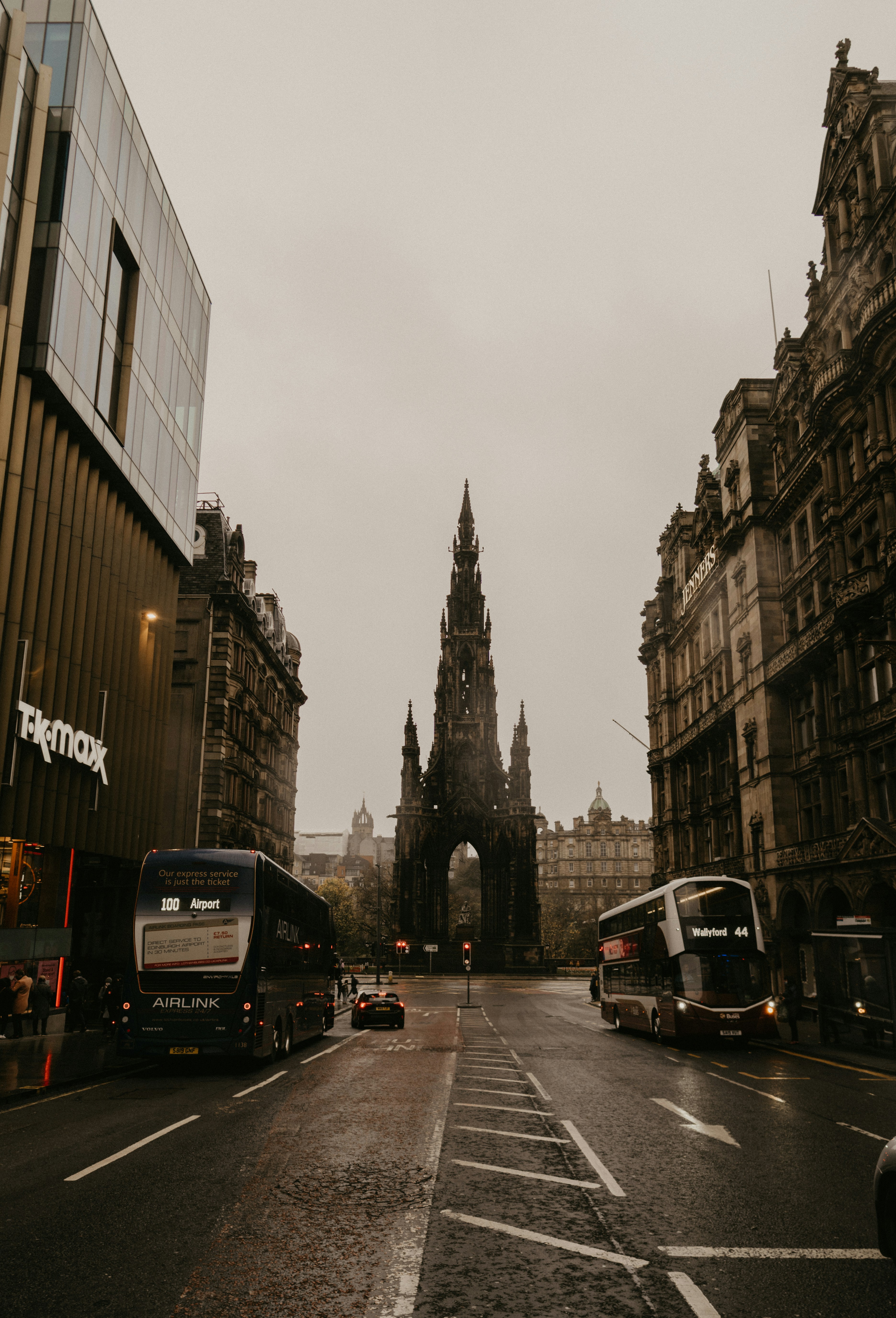 The Scott Monument rises dramatically amidst a bustling urban street, flanked by contemporary architecture and city buses. A moody atmosphere envelops the scene.