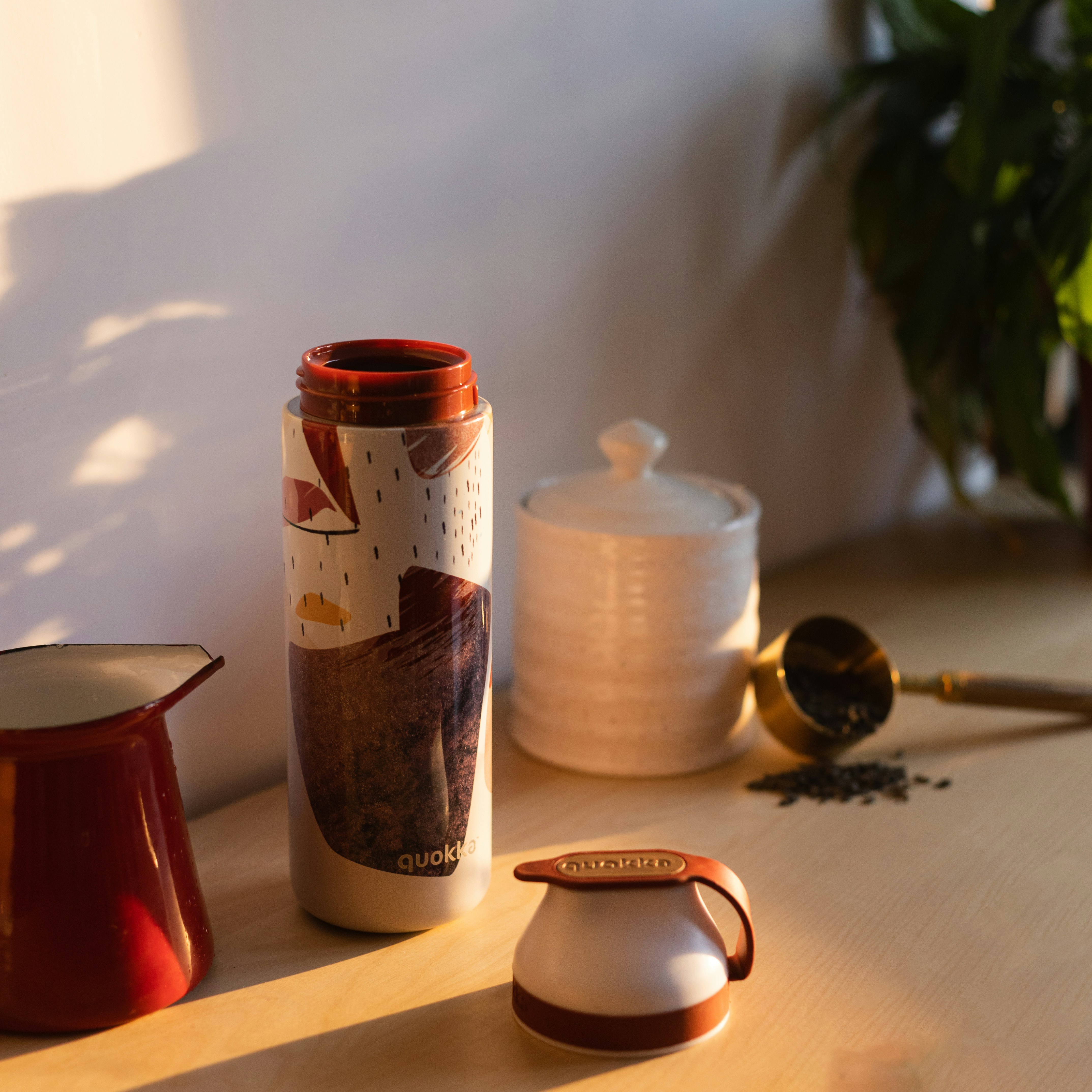 Artistic arrangement featuring a decorated thermos, a white canister, and a measuring spoon on a wooden surface, illuminated by warm sunlight.