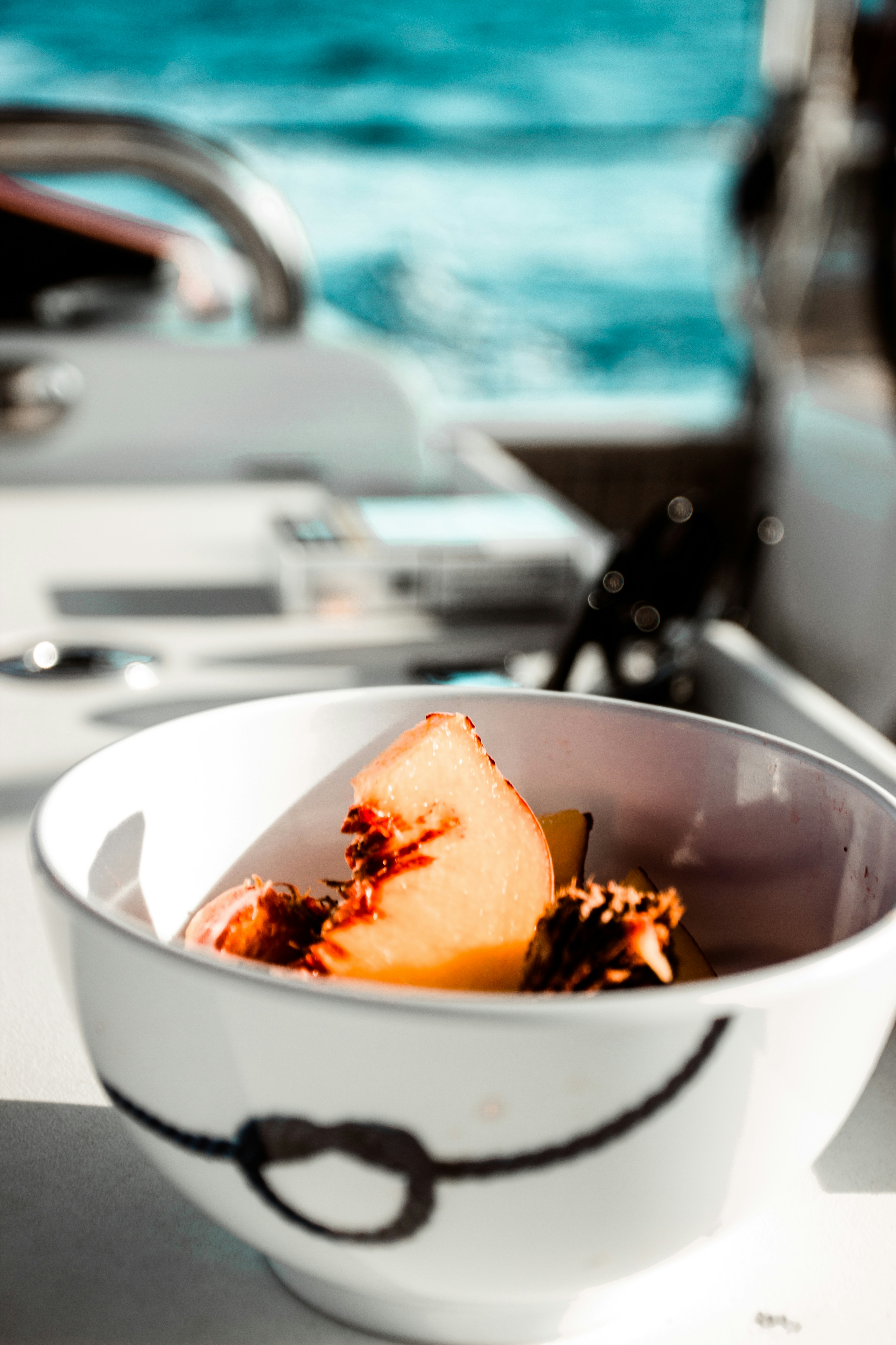 Photograph of a white ceramic bowl with peach slices on a sunlit boat deck, turquoise water blurred in the background.
