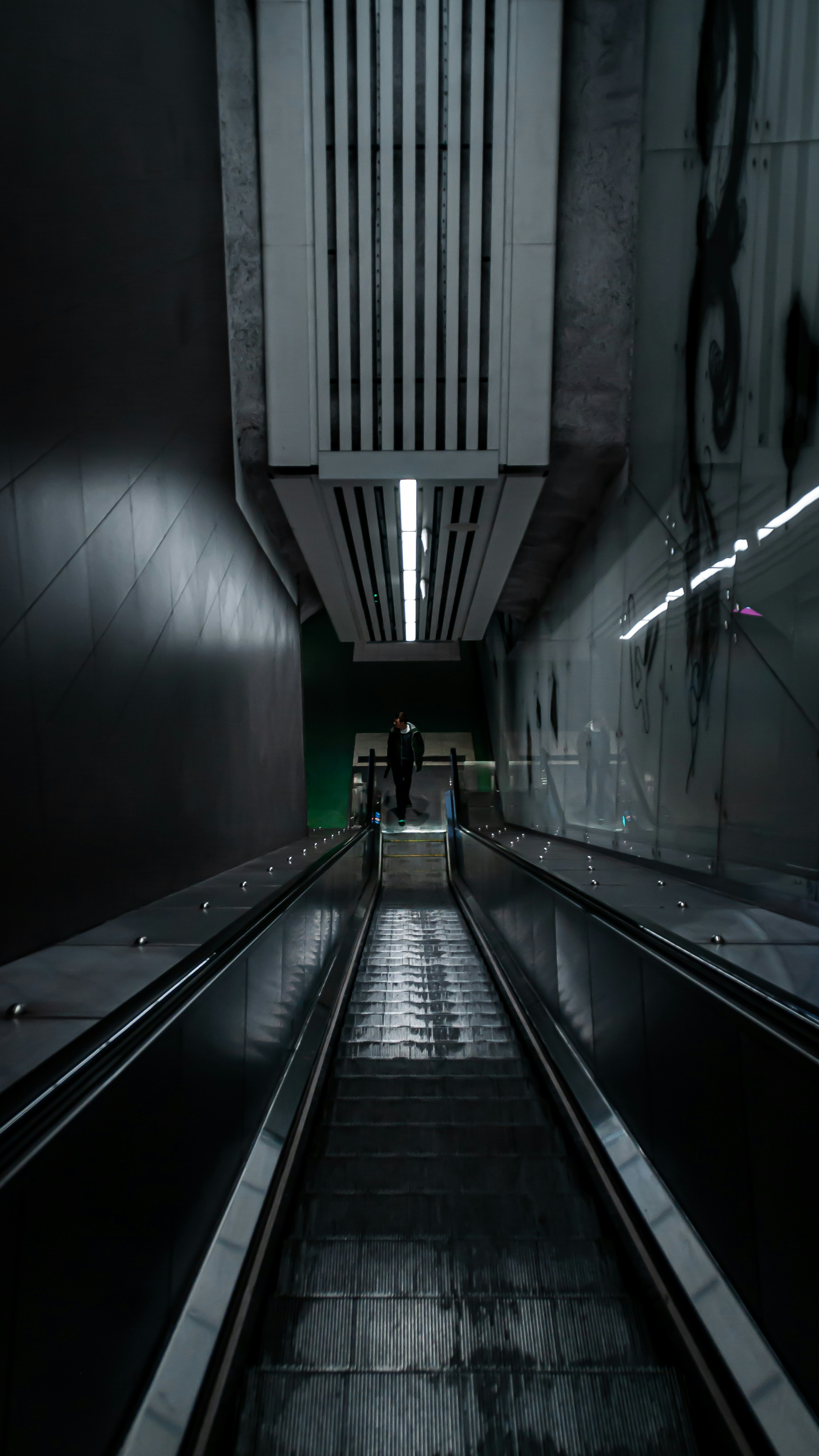 A lone figure descends an escalator in a dimly lit urban space, emphasizing the contrast between light and shadow. The sleek architecture frames the scene.