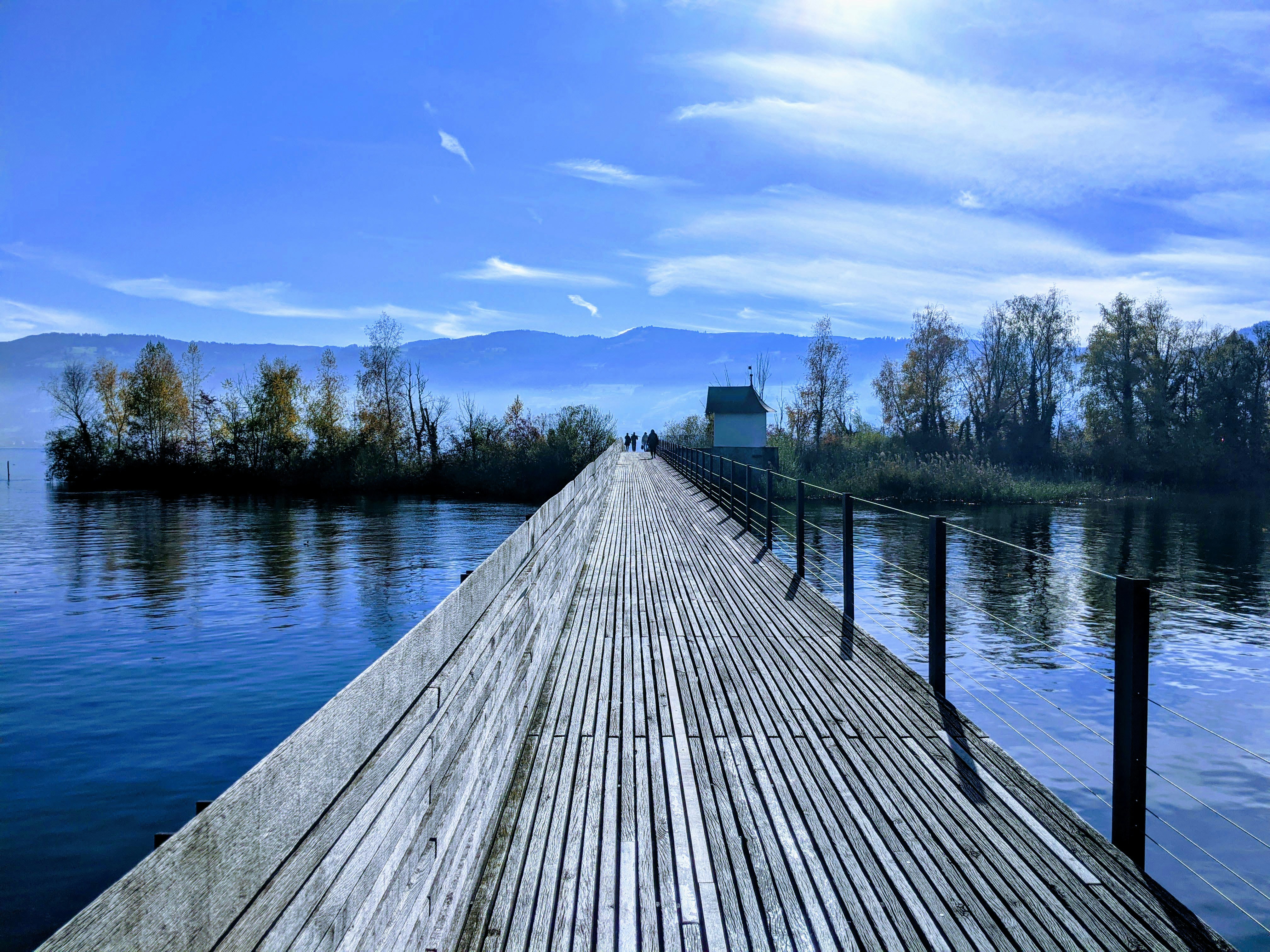 Wooden pier extending into a serene lake, framed by autumn trees and distant mountains. The scene conveys a peaceful, reflective atmosphere.
