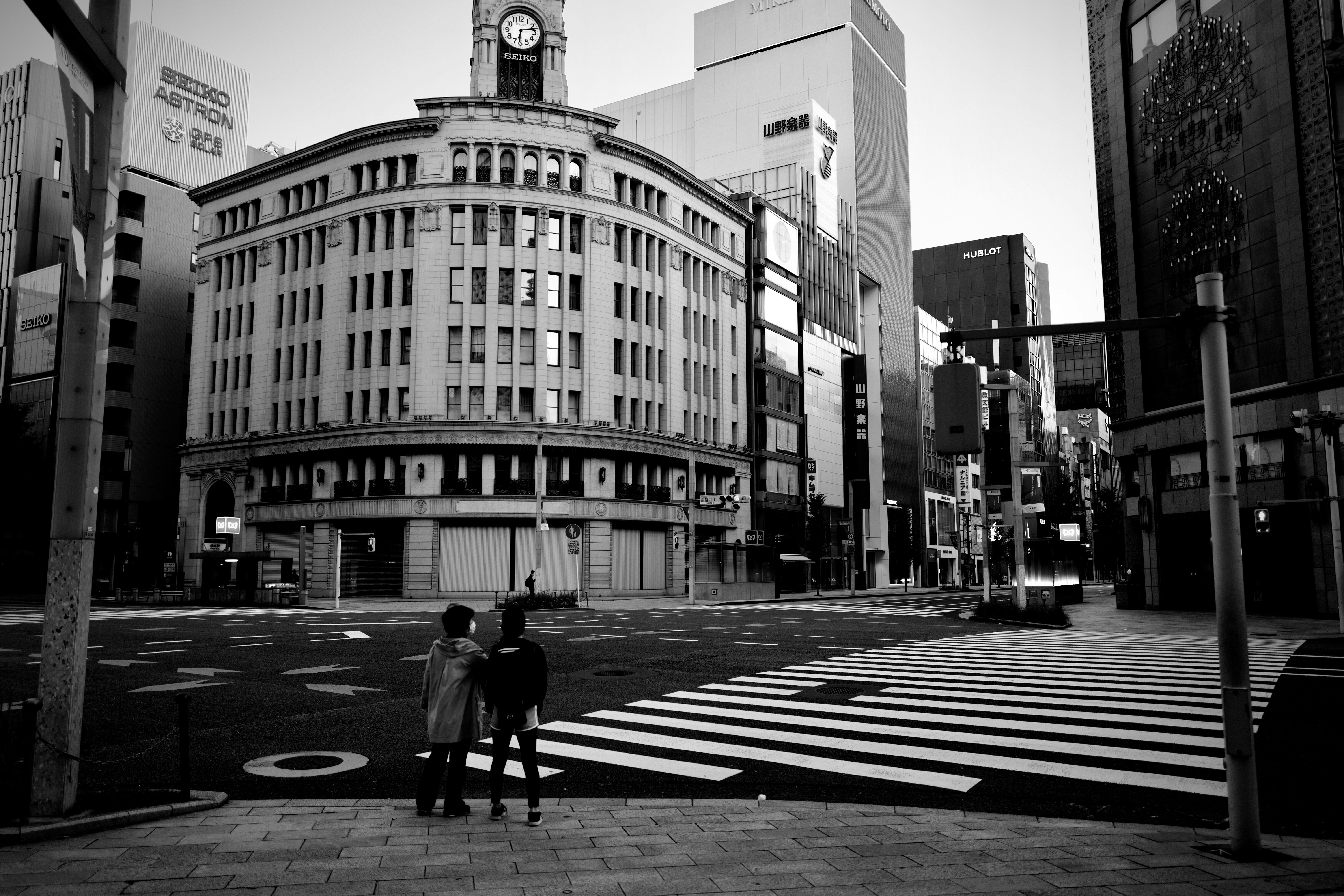 Monochrome cityscape with a couple crossing a quiet intersection against a backdrop of towering buildings.