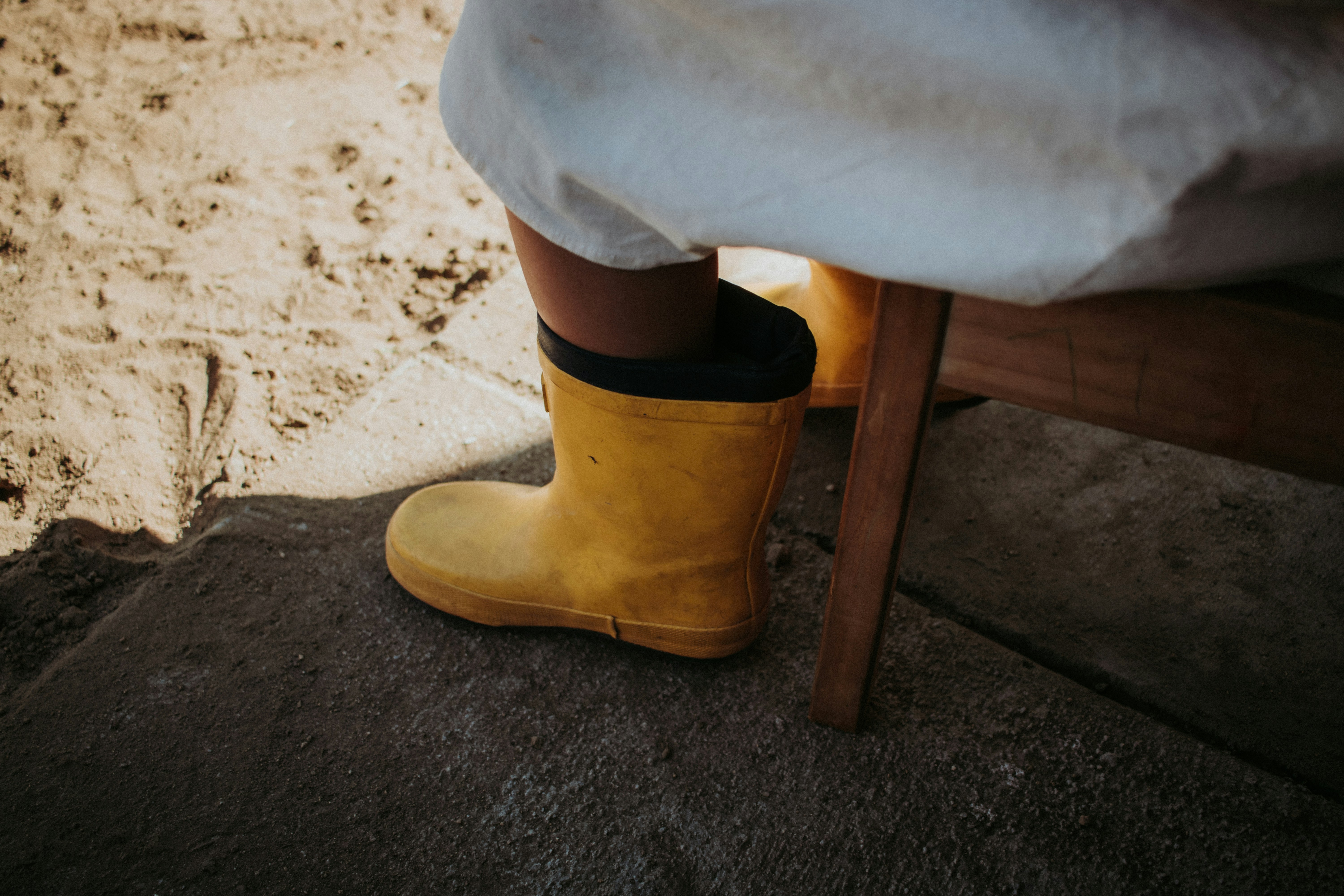 Yellow rubber boots resting beside a wooden chair on a sandy surface, suggesting a relaxed outdoor setting.