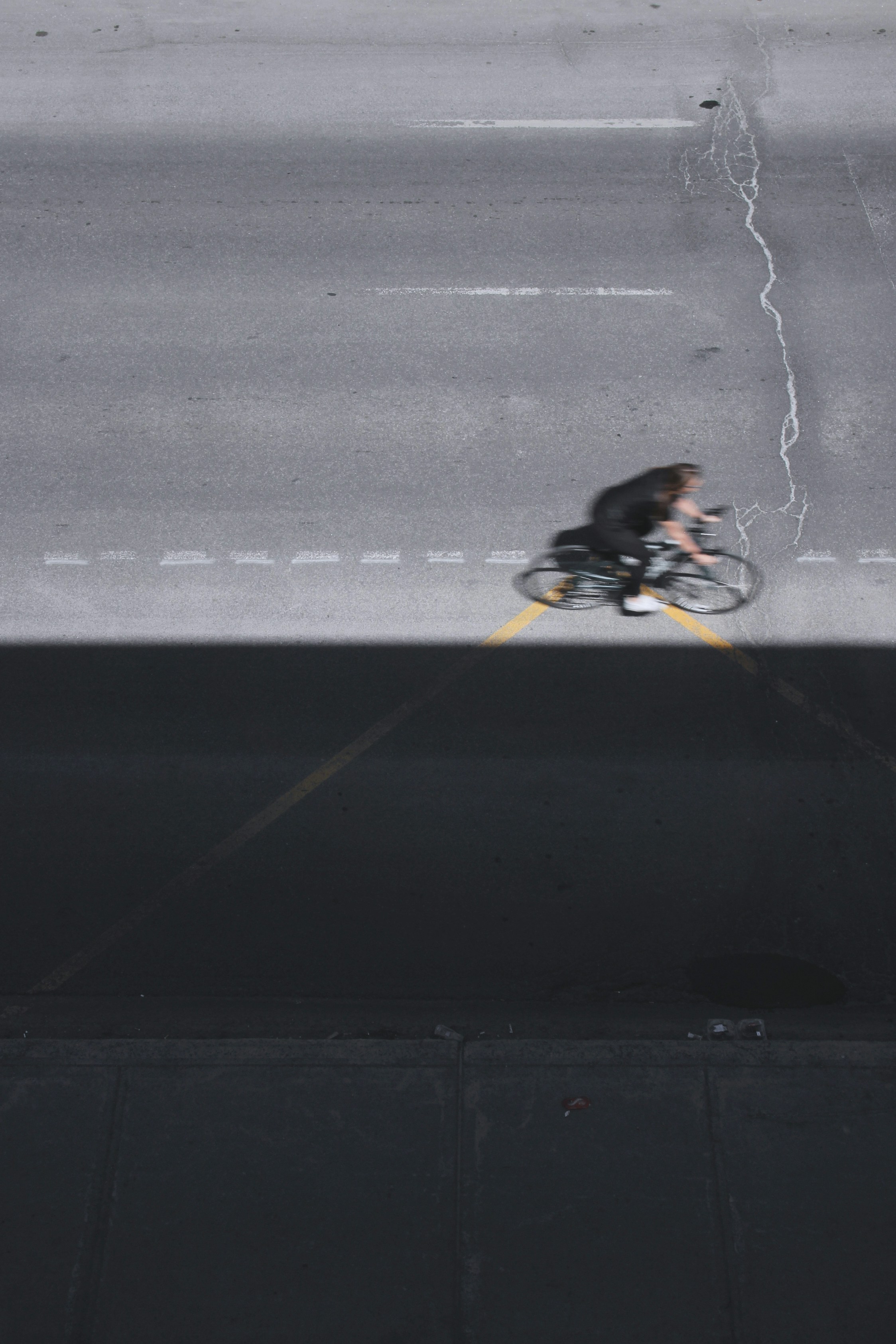 A cyclist rides swiftly across a sunlit street, casting a shadow that contrasts with the asphalt. The dynamic motion captures the essence of urban life.