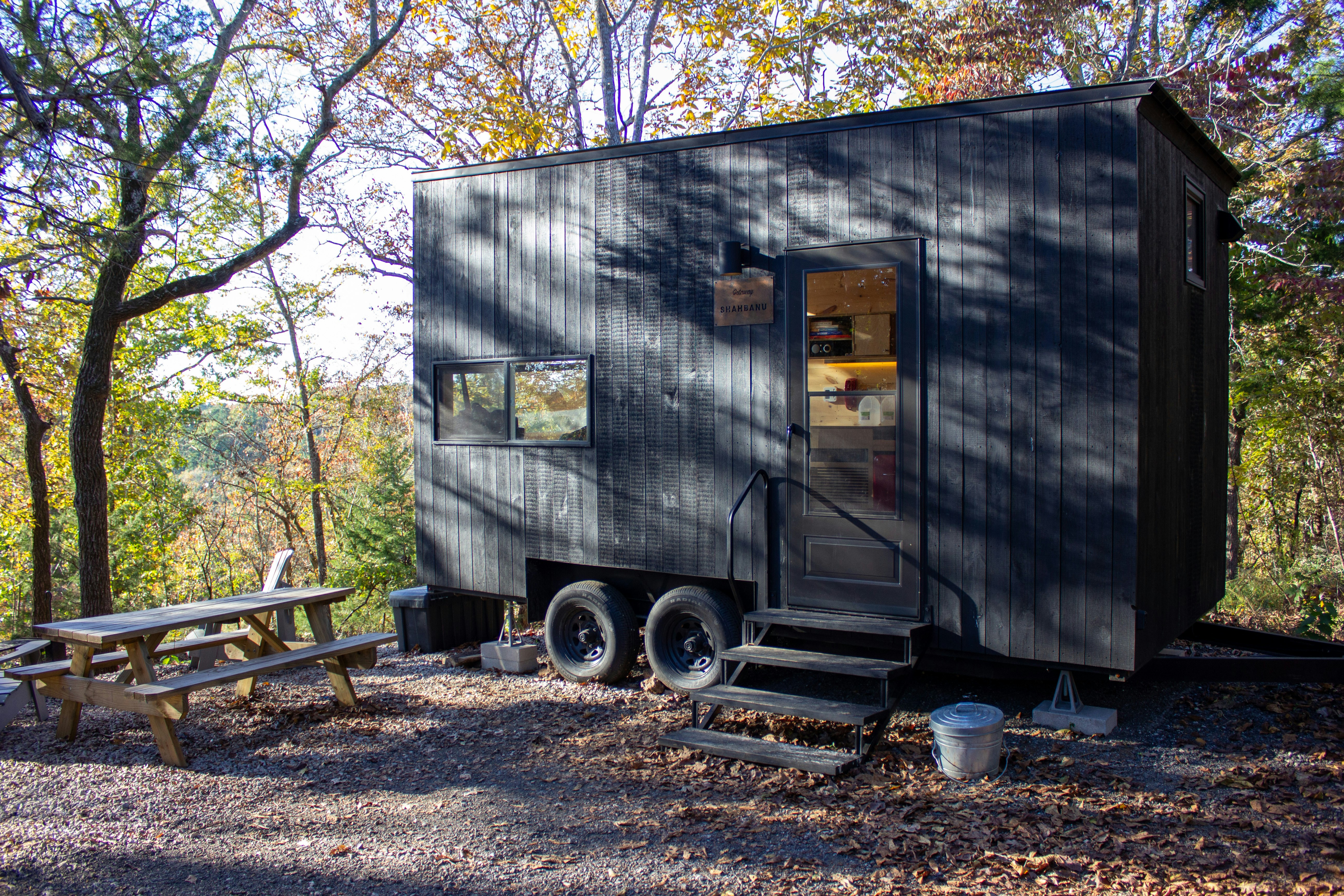 A sleek black tiny house nestled among autumn trees, featuring a simple staircase and outdoor seating area. The structure's modern design contrasts with the natural surroundings.