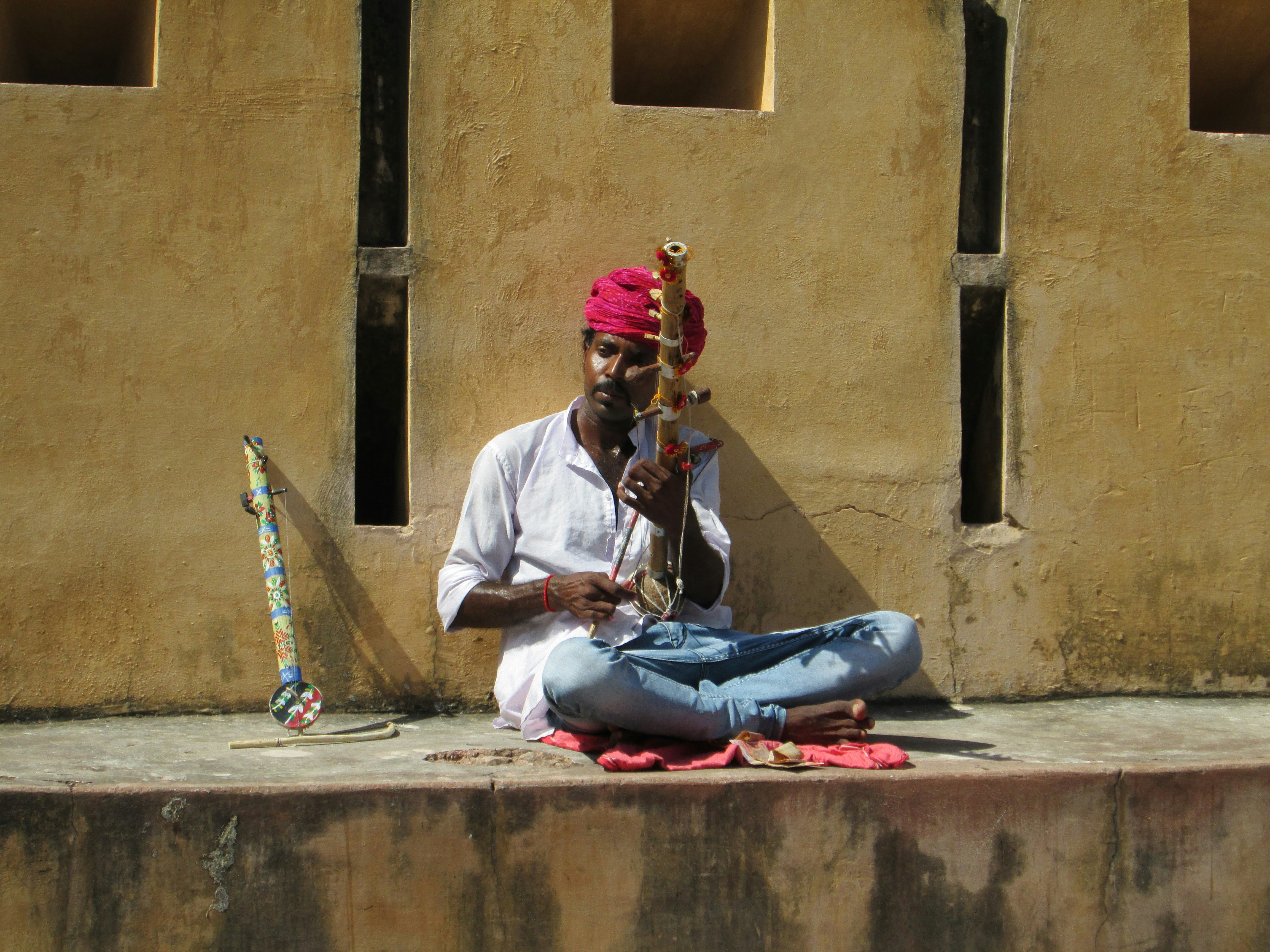 An Indian street musician in a white shirt and red turban sits cross-legged on a sunlit ledge, playing a long traditional reed instrument.