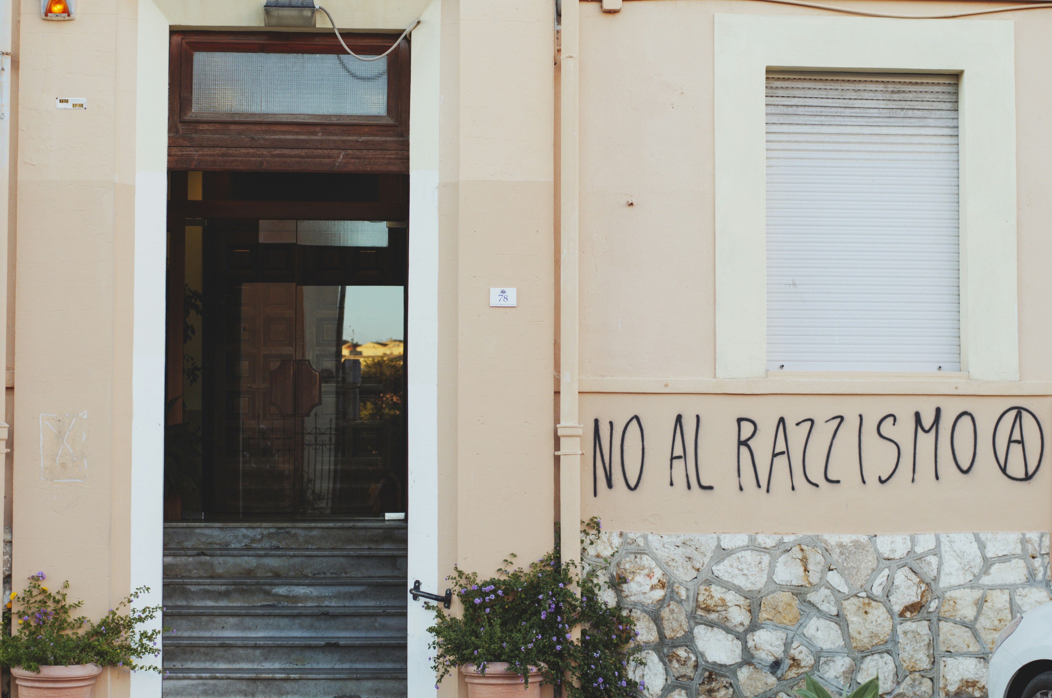 Graffiti reading 'NO AL RAZZISMO' adorns the wall beside an entrance, highlighting a message of unity and resistance. The setting features a quaint building with a stone base and potted plants.