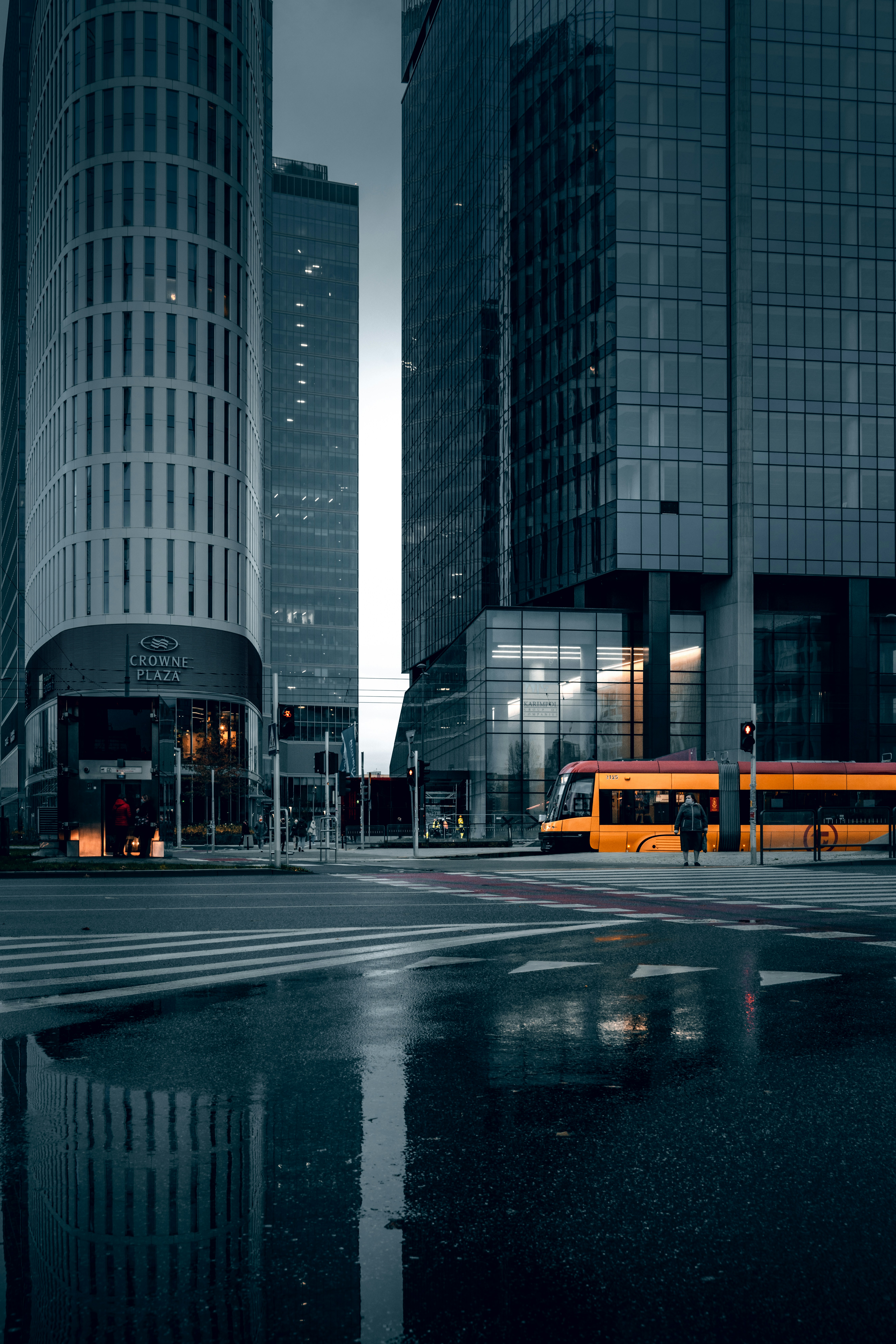 A vibrant orange tram navigates through a modern cityscape, its reflection shimmering on a rain-soaked street. Towering glass buildings frame the scene, creating a striking contrast.