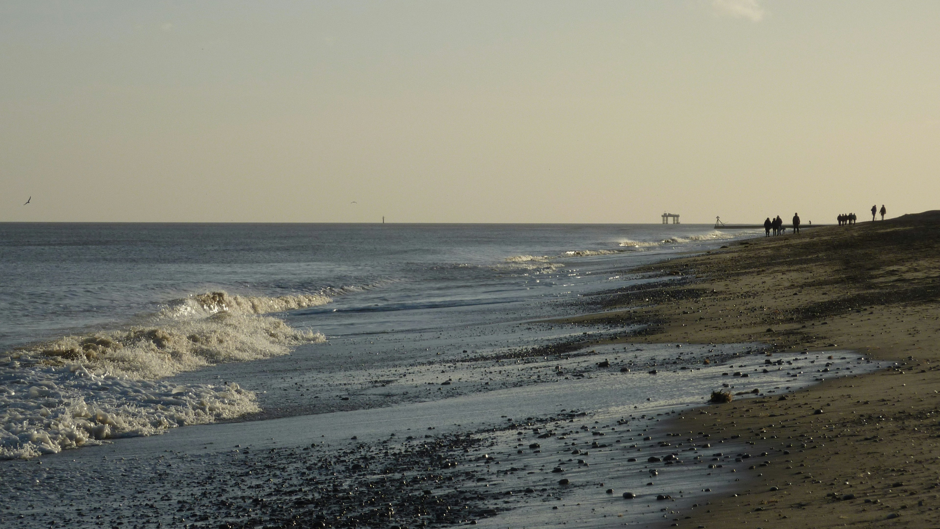 Gentle waves lap against a sandy shore, with silhouettes of people strolling along the beach. The sun sets, casting a soft glow over the tranquil scene.