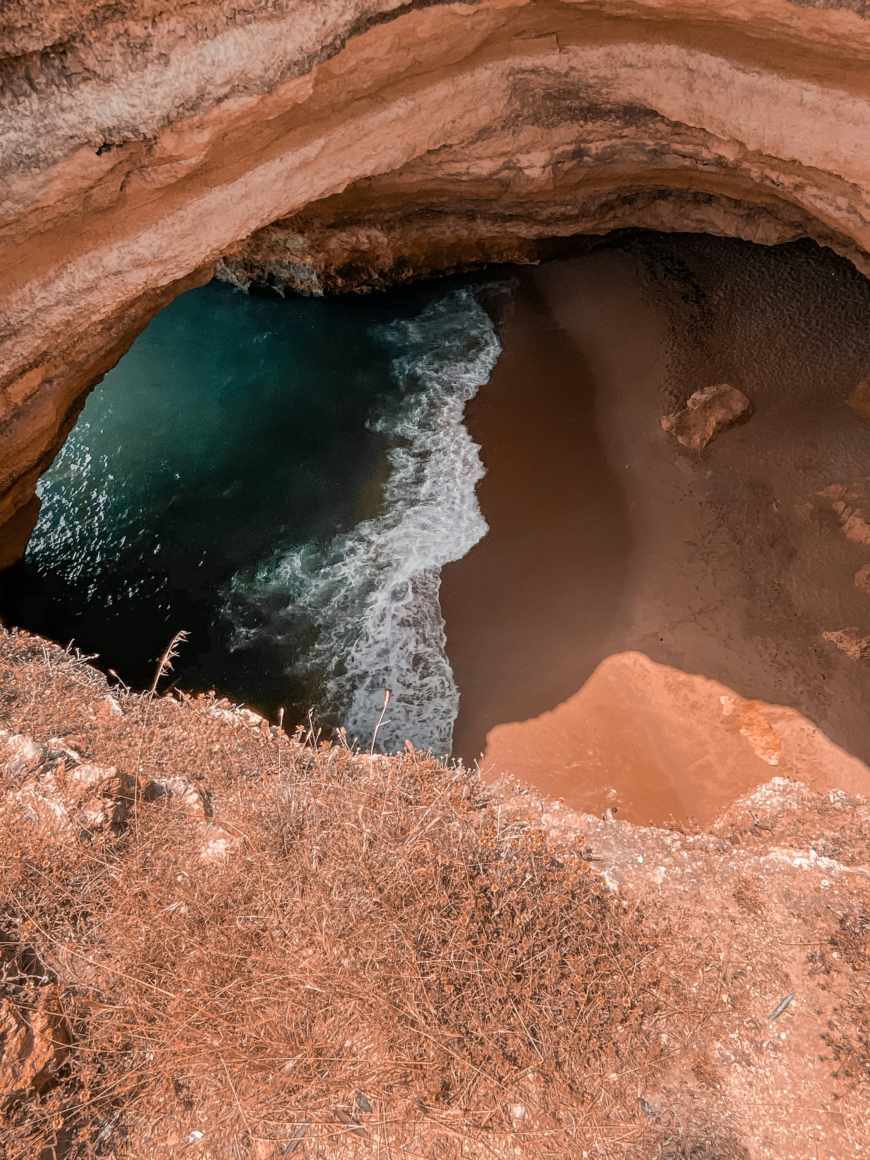 Aerial view of a secluded cove where turquoise waters meet sandy shores, framed by rugged cliffs and dry vegetation.