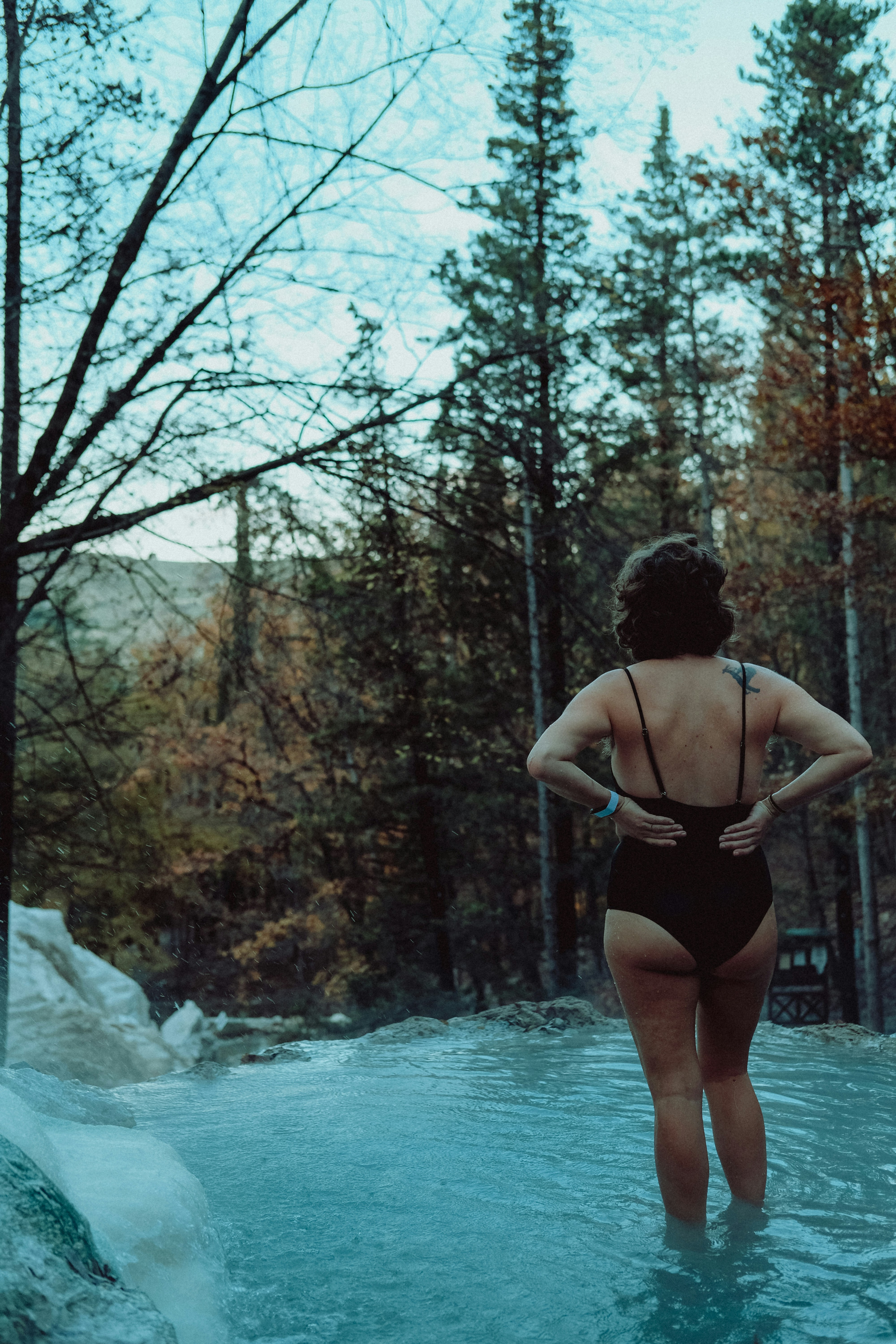 Woman in black swimsuit standing in a pool surrounded by tall trees.