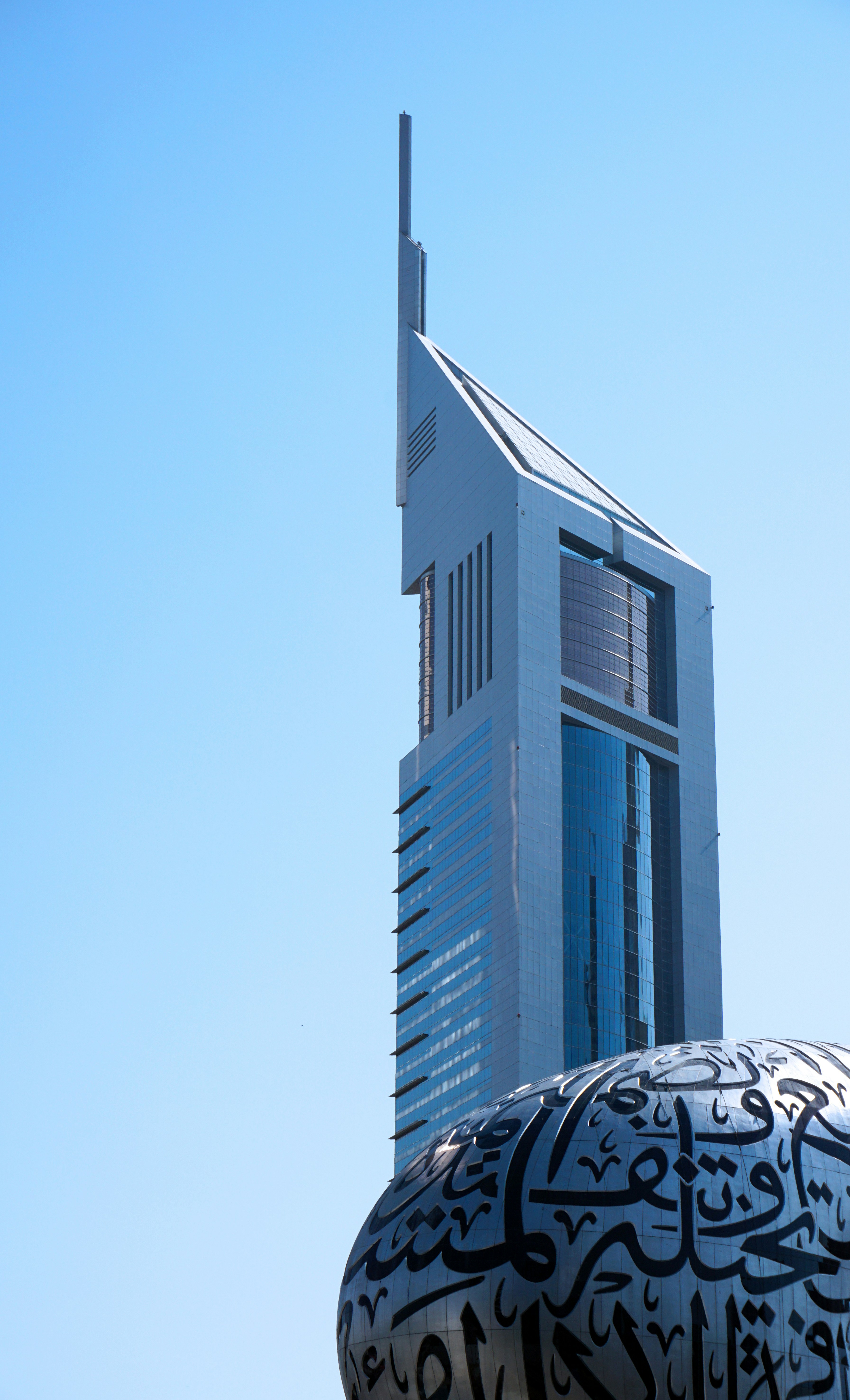 A sleek modern skyscraper rises above an intricately designed dome featuring Arabic calligraphy, showcasing a blend of contemporary architecture and cultural heritage.