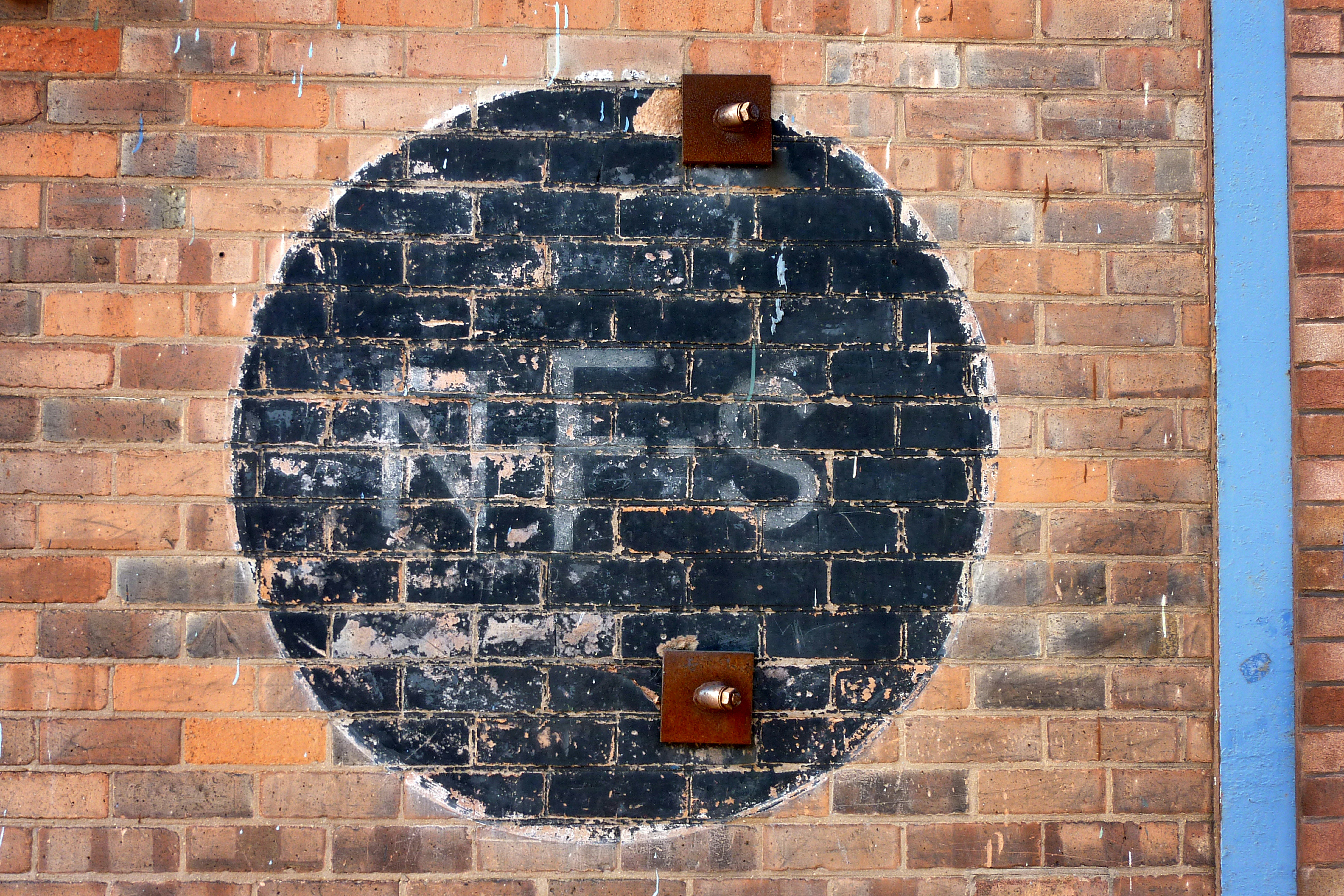 Dark circular patch on a brick wall with two rusted square brackets; a vertical blue metal stripe on the right. The circle appears painted or weathered, revealing a faded emblem.
