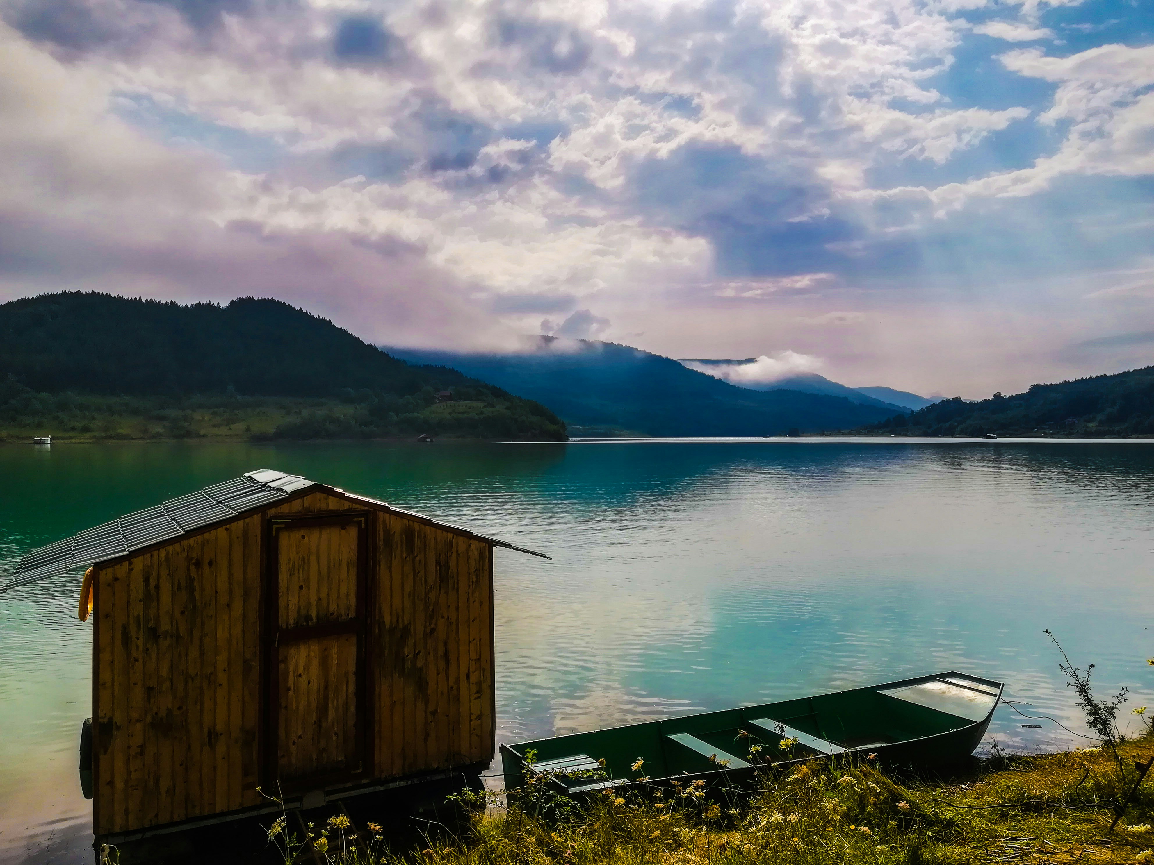 Wooden cabin and boat by a tranquil lake under a cloud-dotted sky.