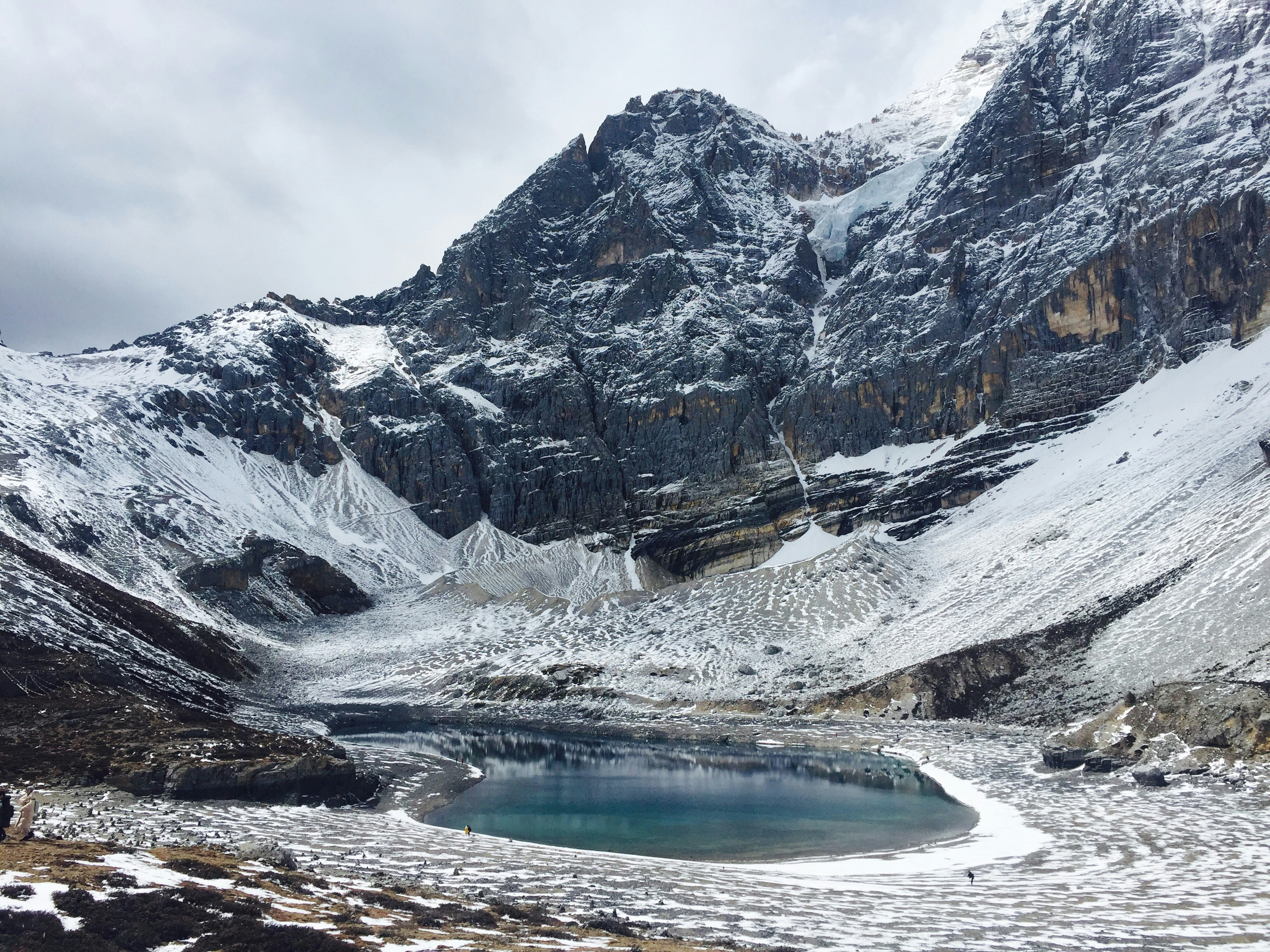 Snow-dusted mountains encircle a tranquil, blue-hued lake under a cloudy sky.
