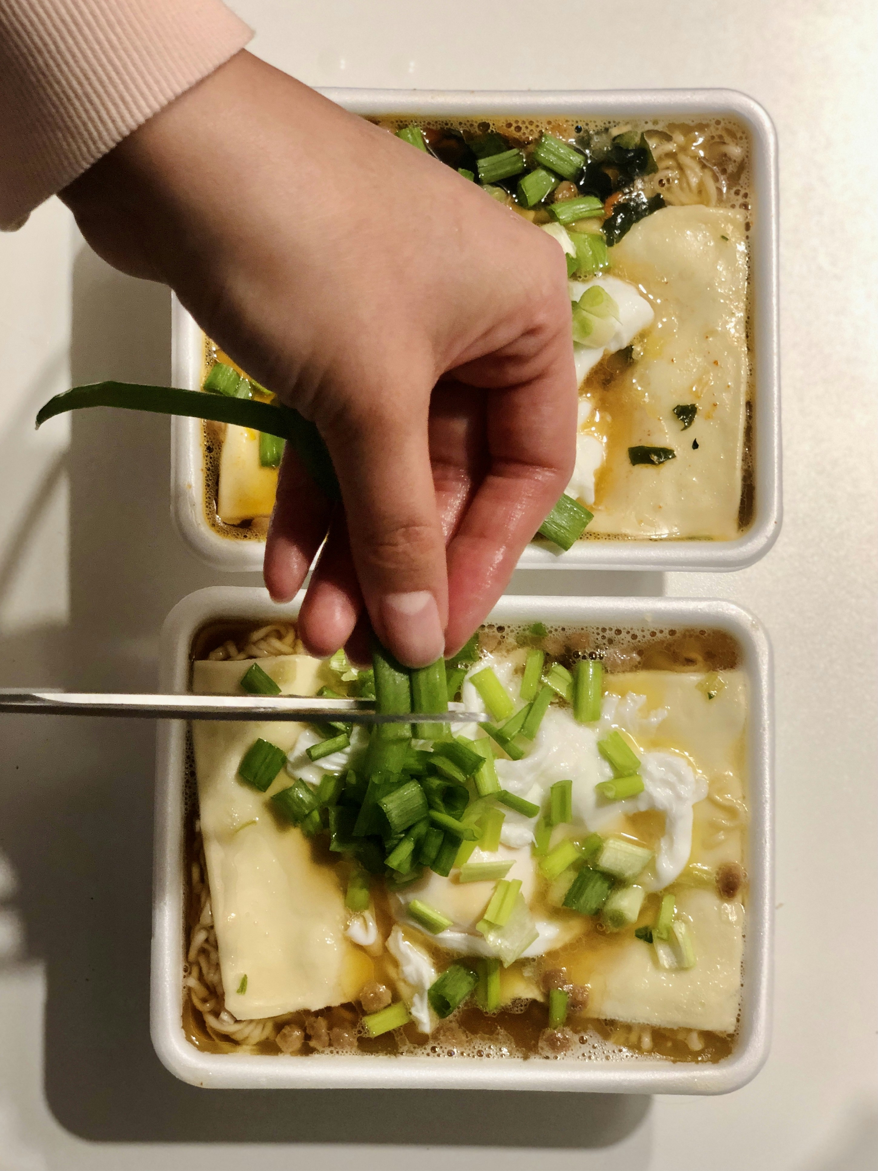 Hand sprinkling chopped green onions over steaming bowls of ramen with cheese and egg. The image highlights the final touch of flavor enhancement.