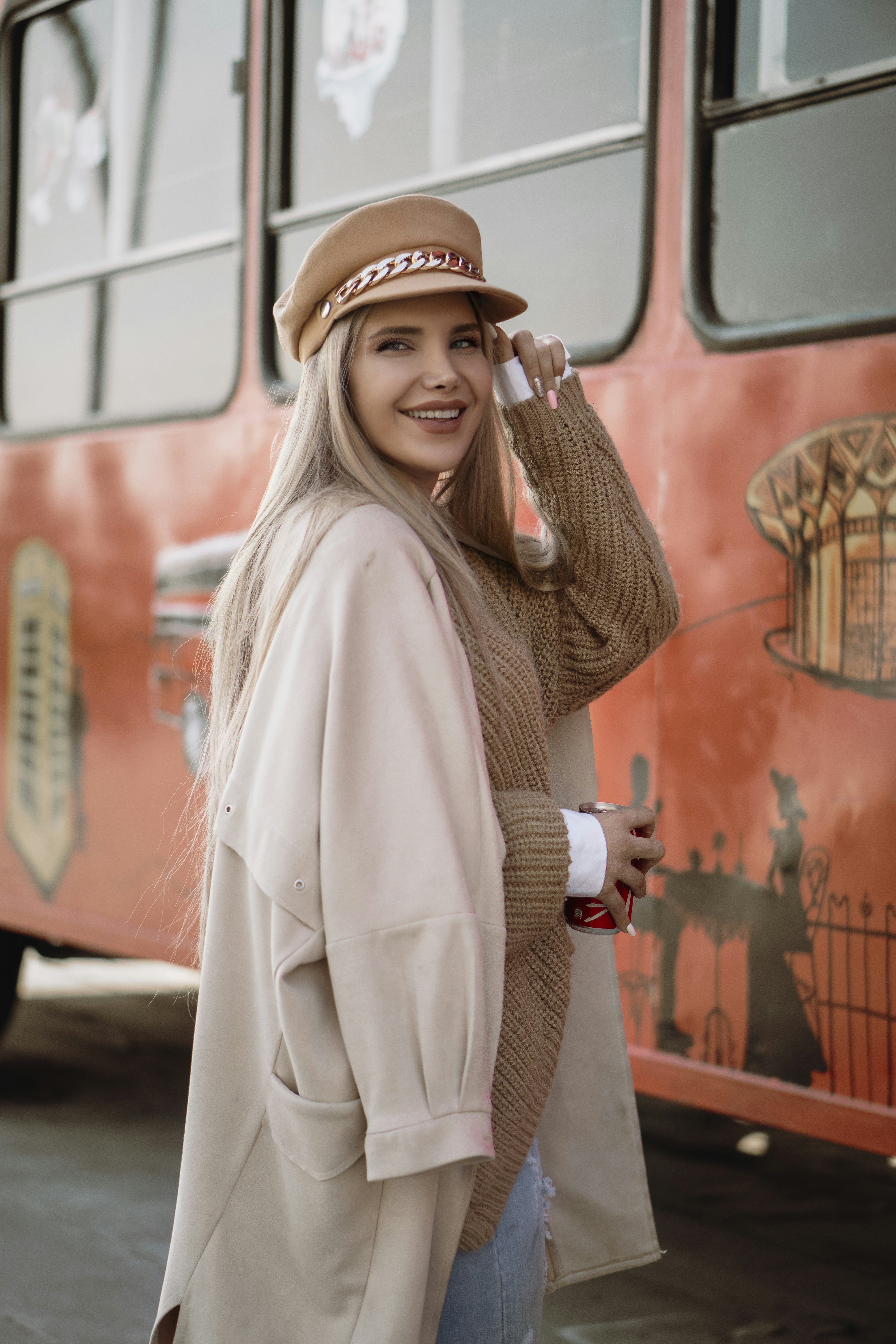 Young woman smiling and adjusting her hat in front of a vintage tram, showcasing a blend of contemporary style and nostalgic charm.
