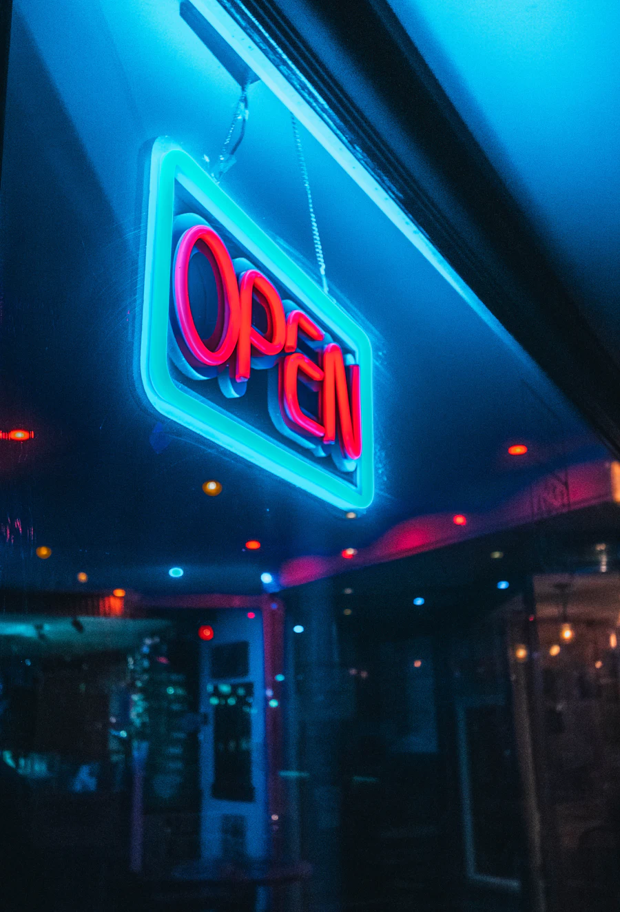 Glowing blue-and-pink neon Open sign hanging inside a dark storefront with colorful reflections and bokeh lights.