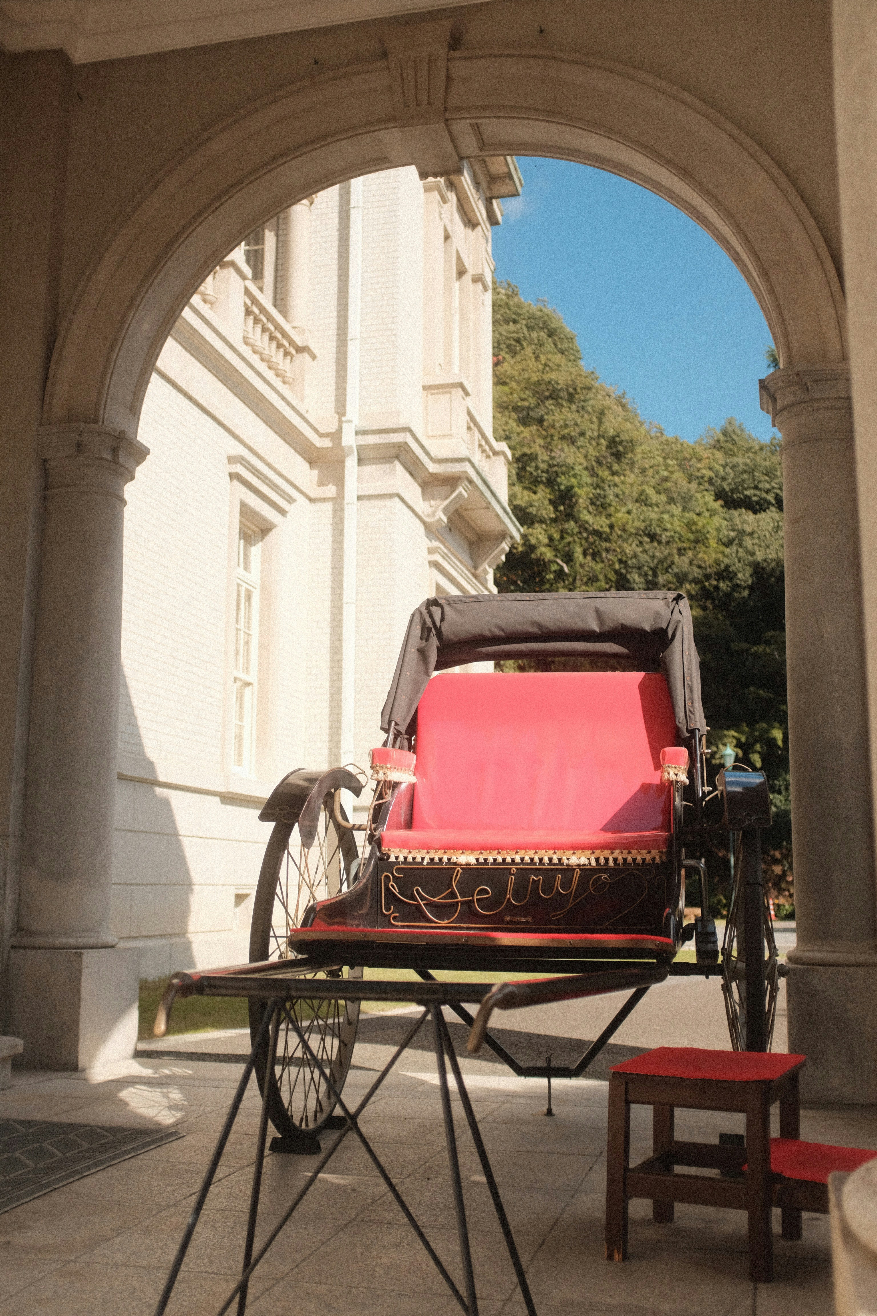 Vintage rickshaw parked under an archway with a sunlit backdrop of lush greenery. The scene evokes nostalgia and charm.