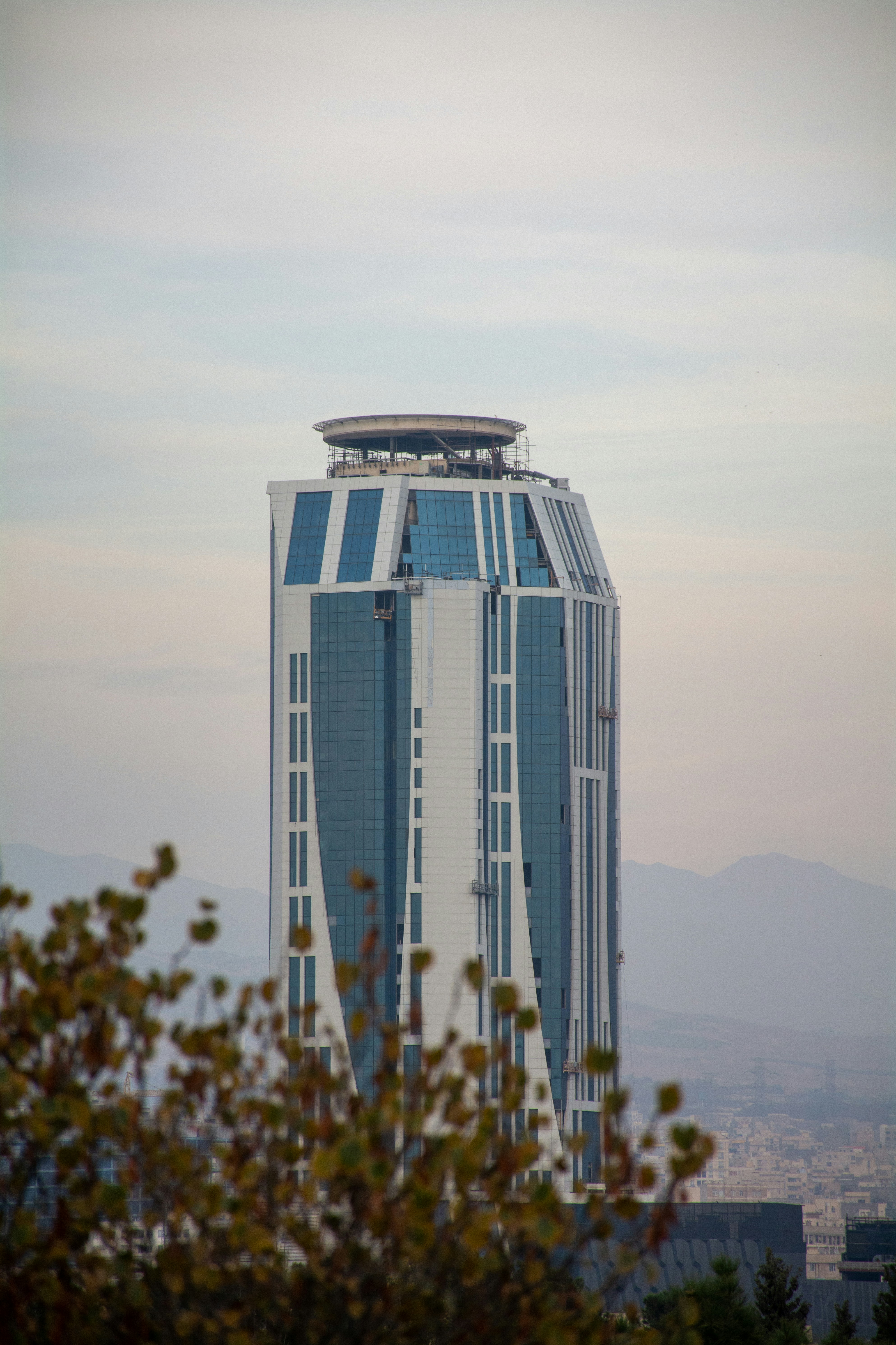 A modern skyscraper with a unique design rises above the cityscape, framed by foliage in the foreground.