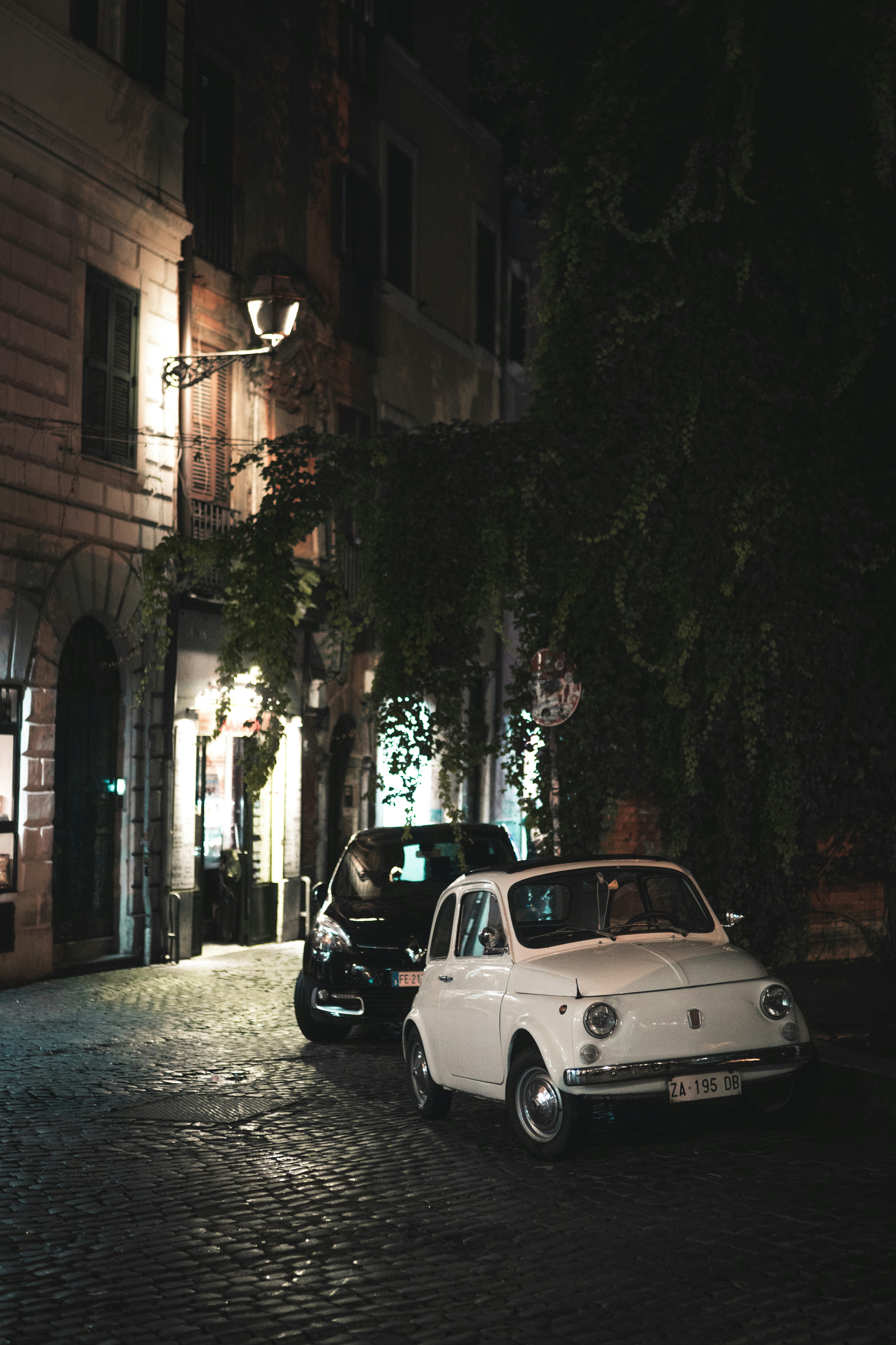 A classic white car parked on a cobblestone street, illuminated by soft streetlights amidst lush greenery and historic buildings.