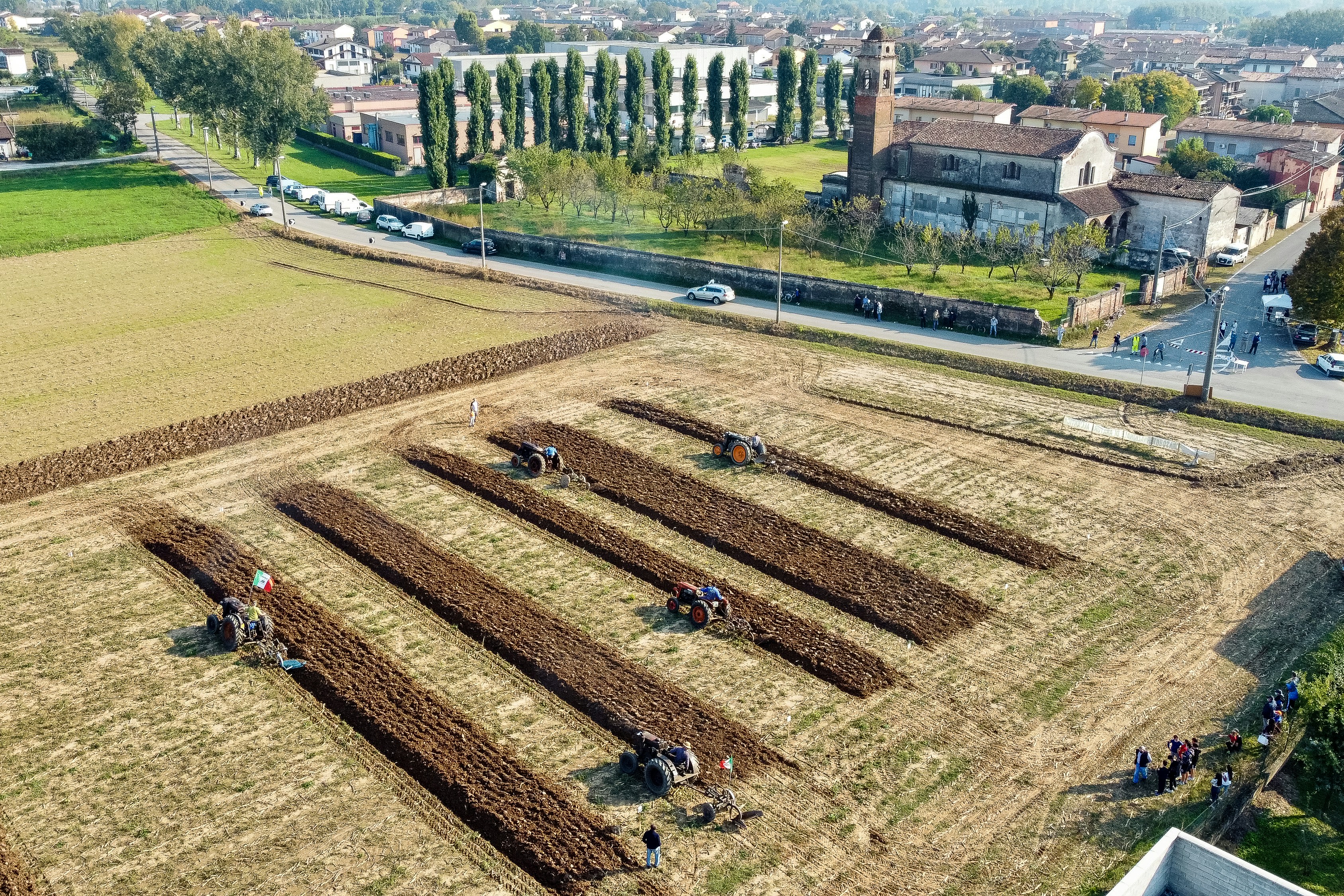 Aerial view of tractors plowing fields in neat rows with a village in the background.