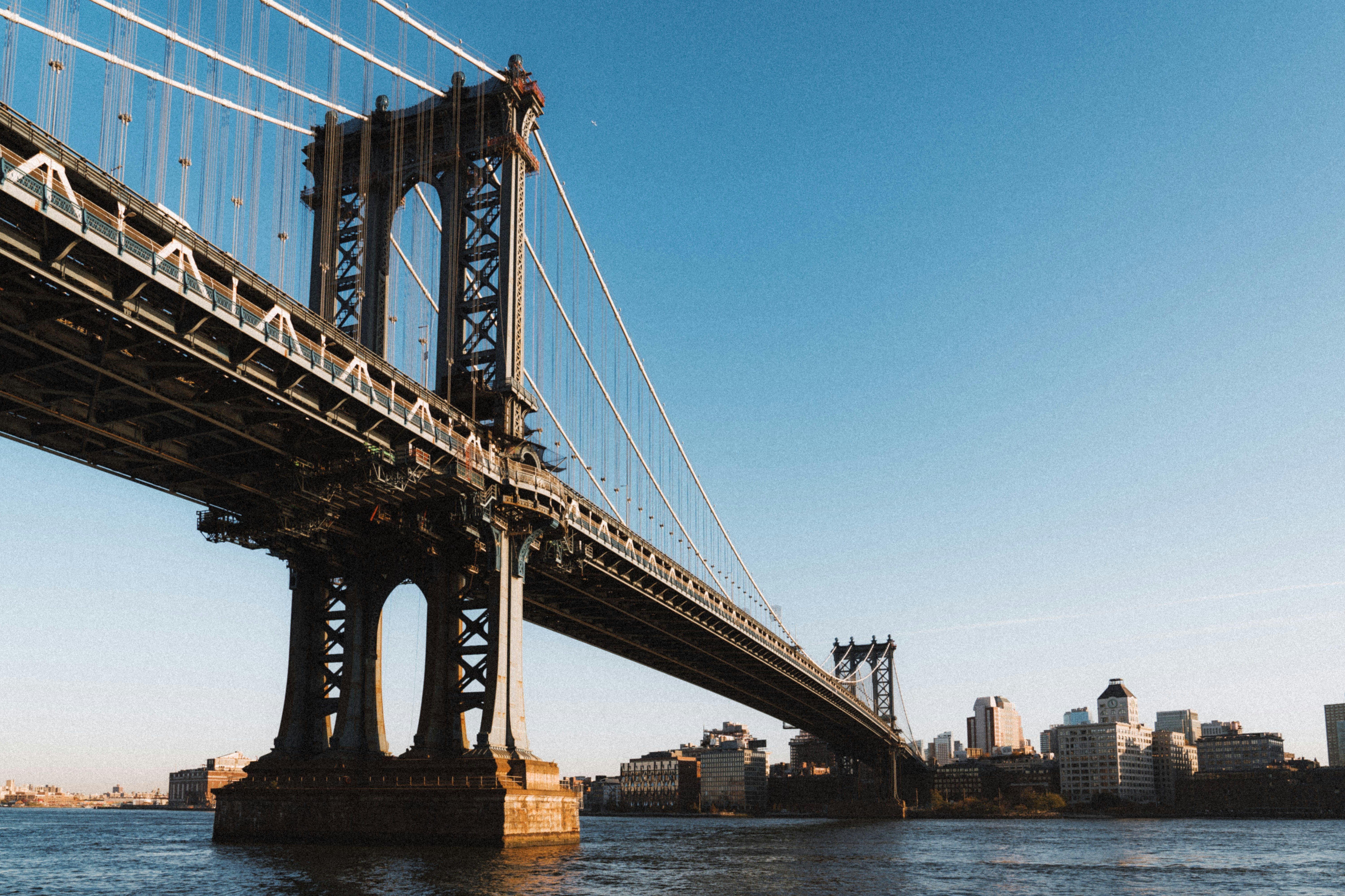 The Manhattan Bridge stretches majestically across the water, showcasing its intricate steel structure against a clear blue sky.
