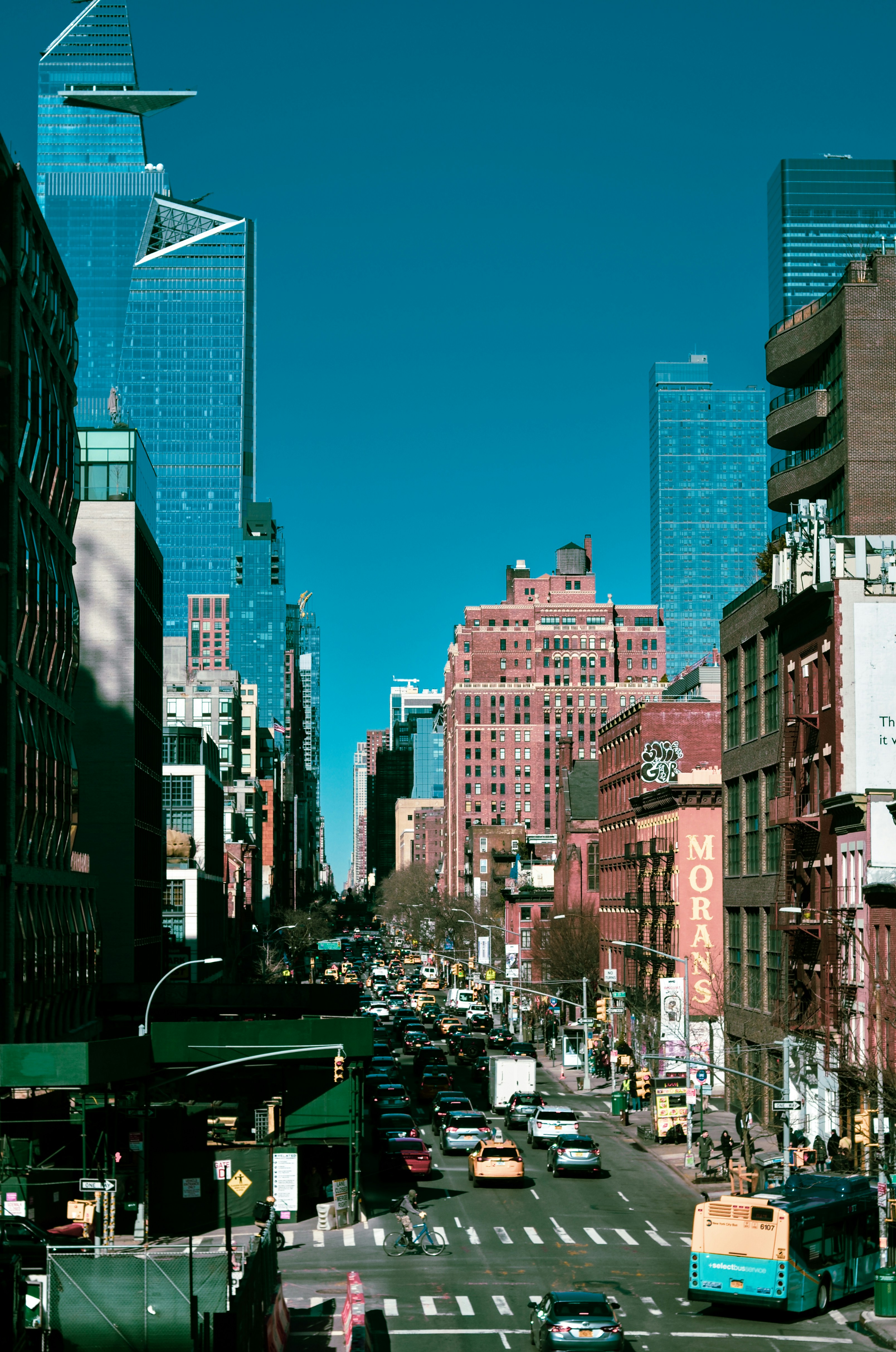 Bustling city street filled with traffic and pedestrians, framed by towering skyscrapers under a clear blue sky.