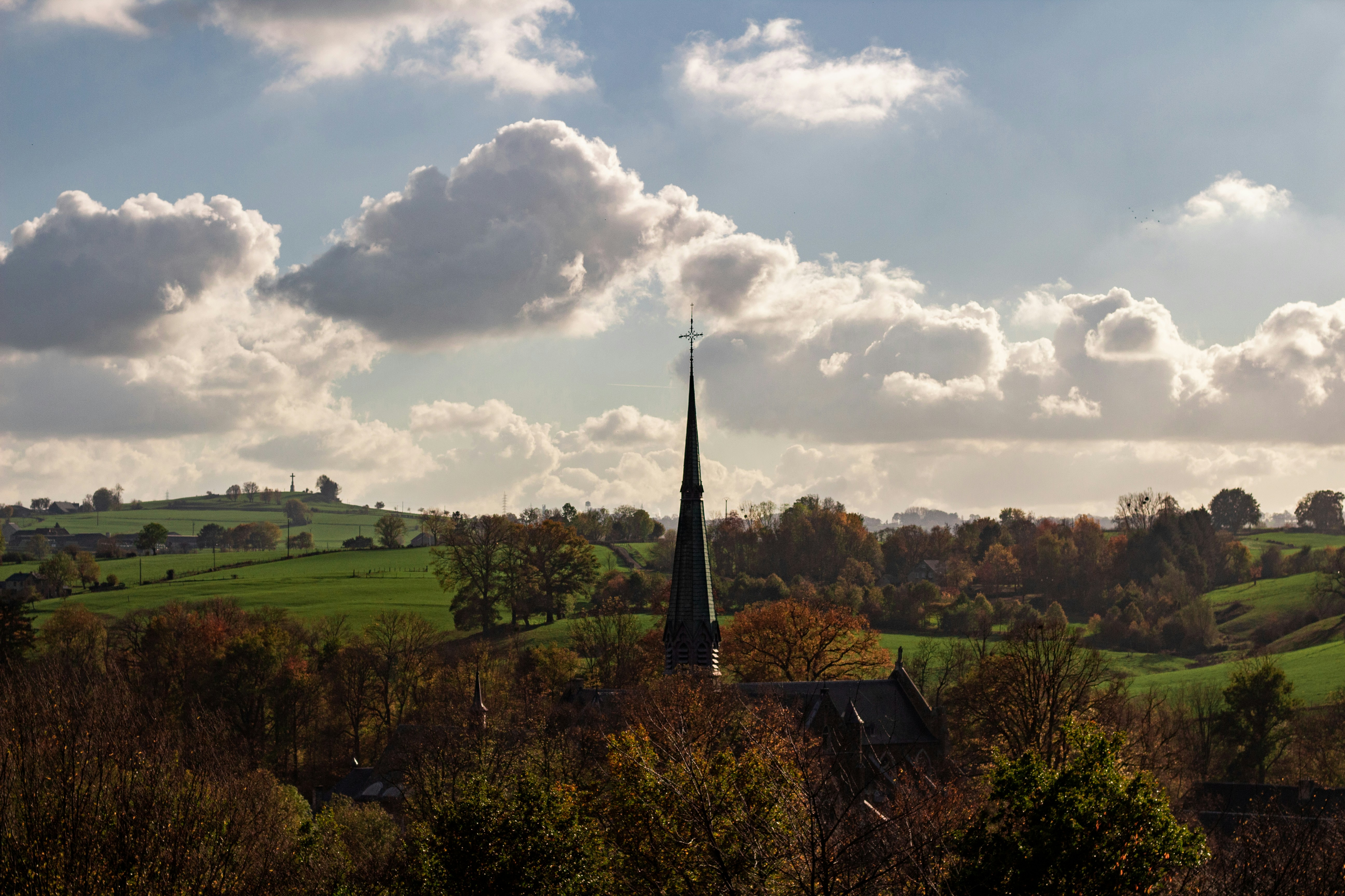 Church steeple rises above vibrant autumn foliage, framed by lush green hills and a dramatic sky filled with clouds.
