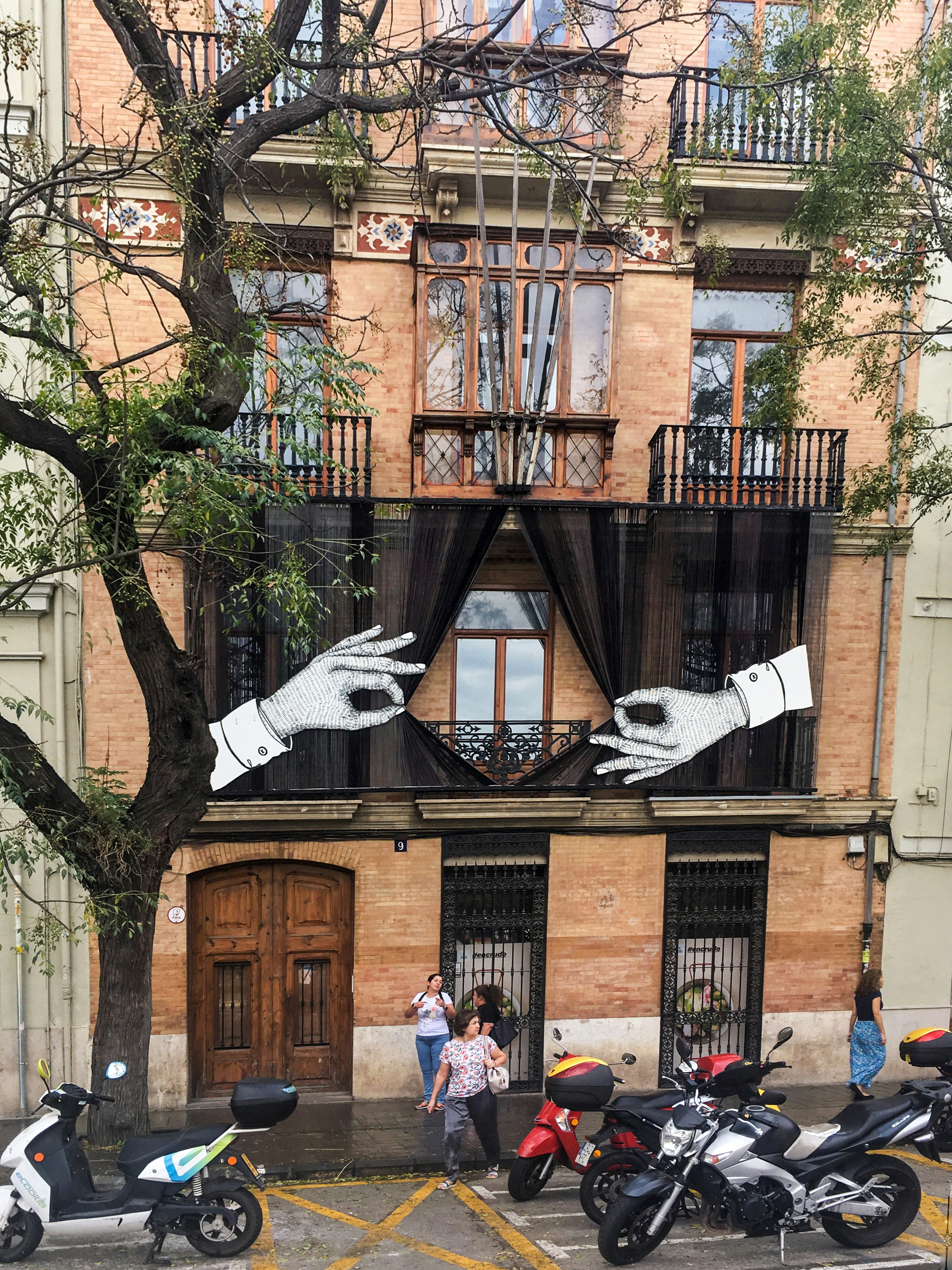 Artistic hands emerge from a balcony draped in black curtains, adding a surreal touch to the historic building's facade. Two pedestrians engage in conversation below.