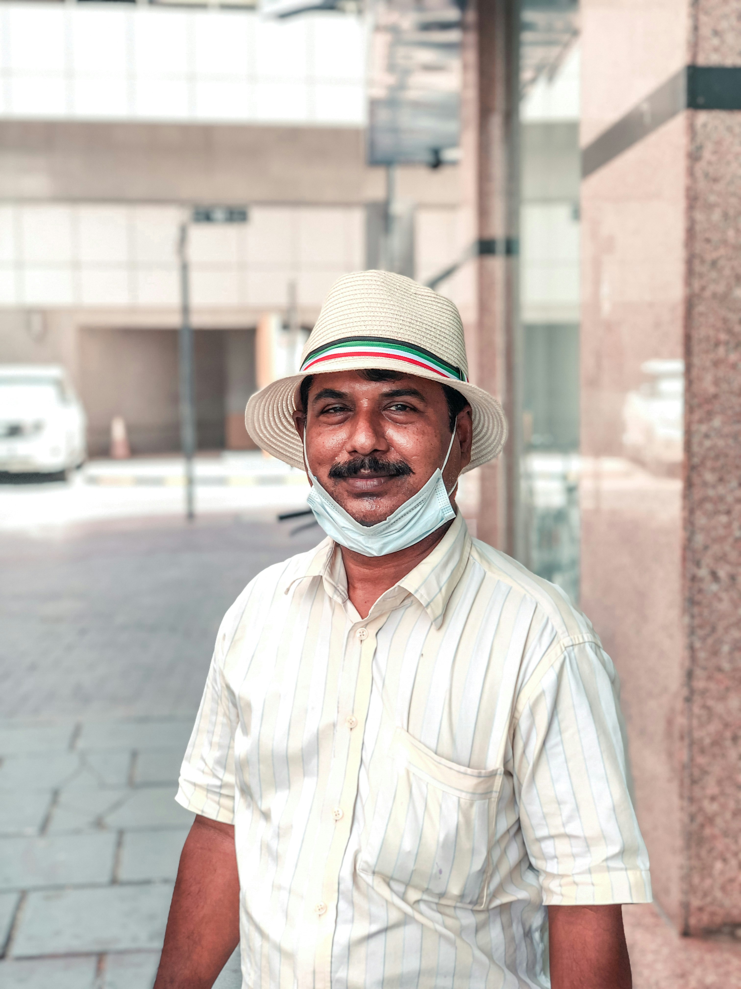 Man wearing a straw hat and a mask stands in an urban setting, exuding warmth and friendliness. The background features a modern architectural facade.