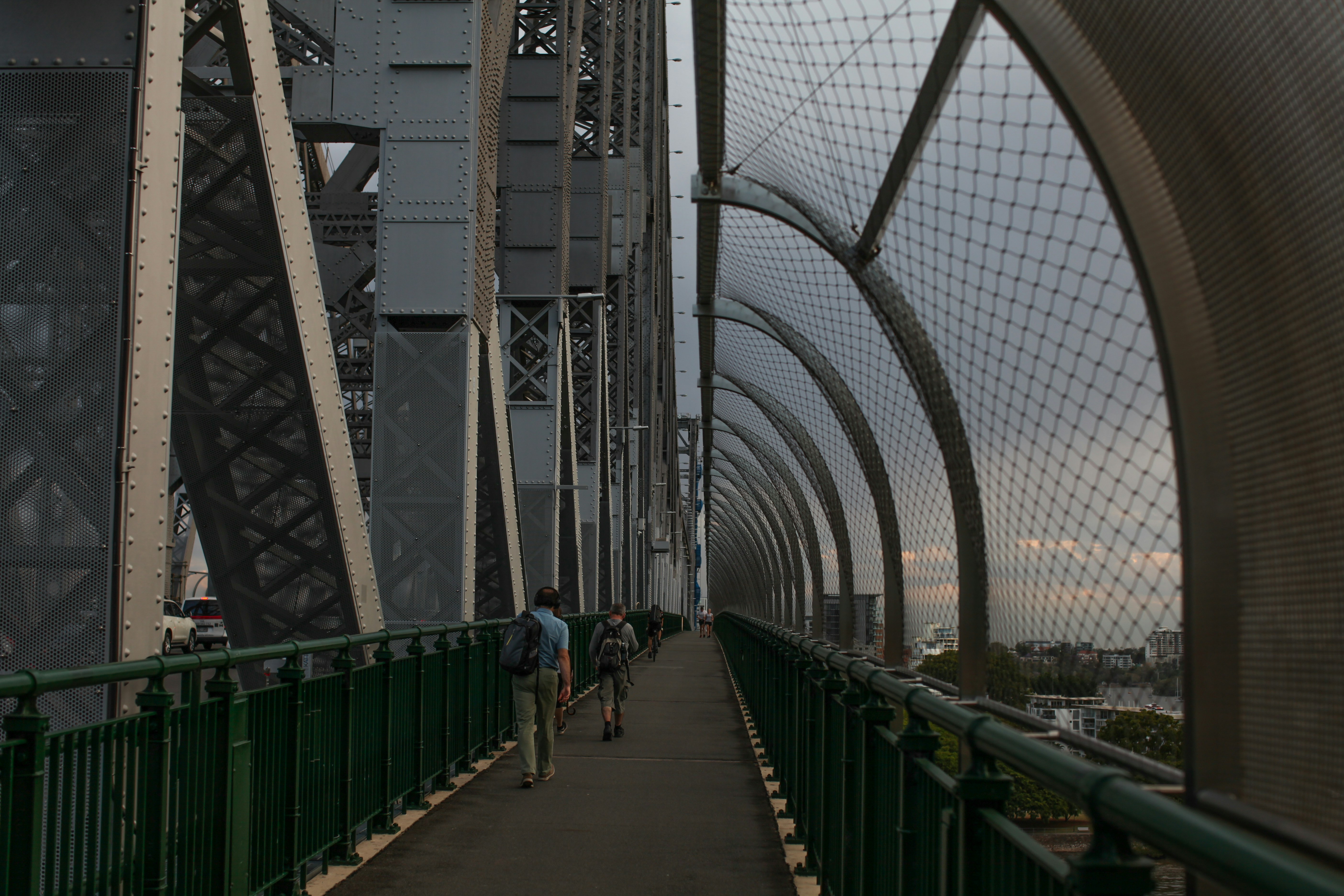 Pedestrians walking along a bridge walkway, framed by intricate steel structures and protective netting.