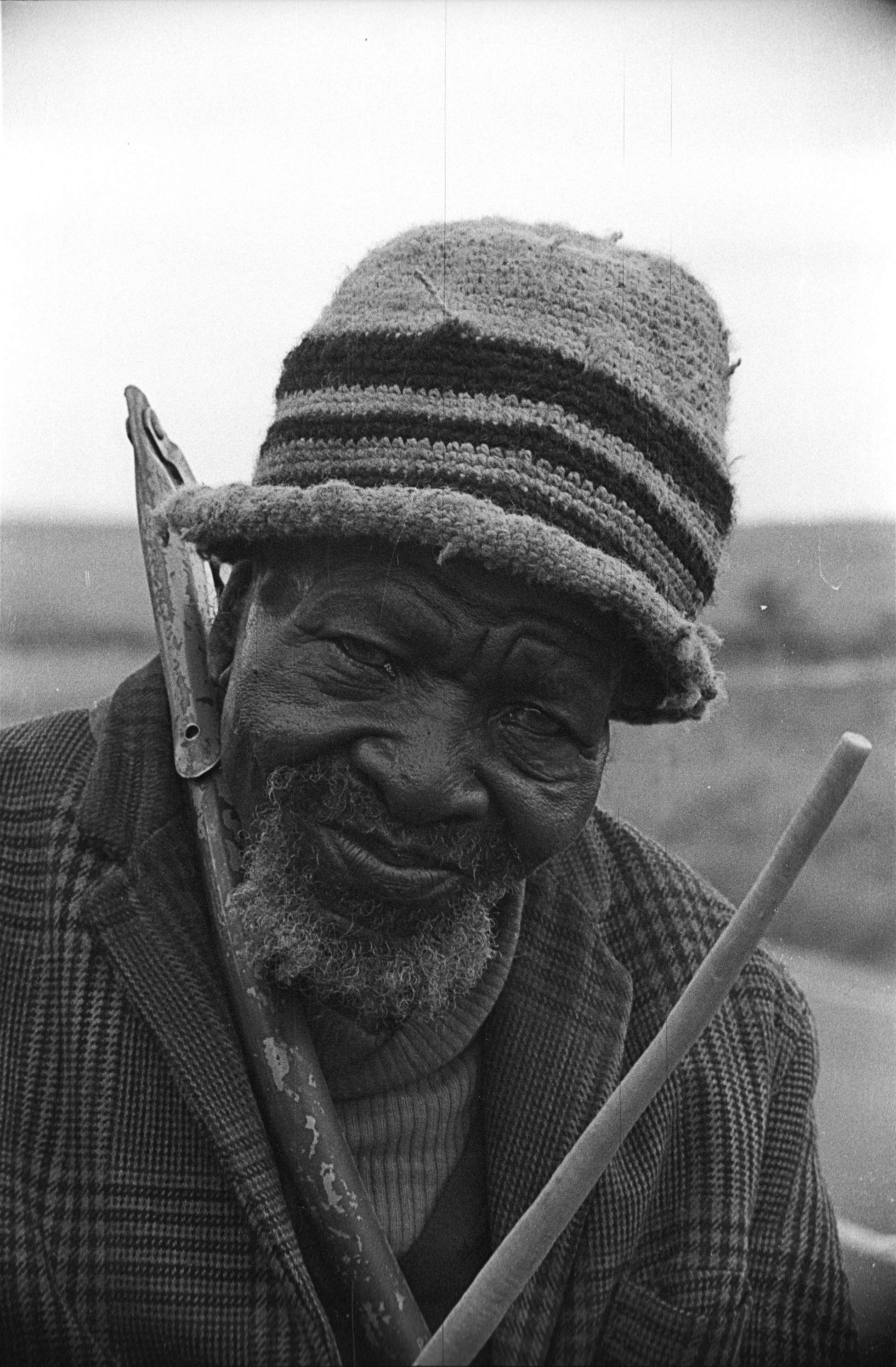 Elderly man with a hand-knit hat and a weathered expression, holding a stick, set against a blurred background of open land.