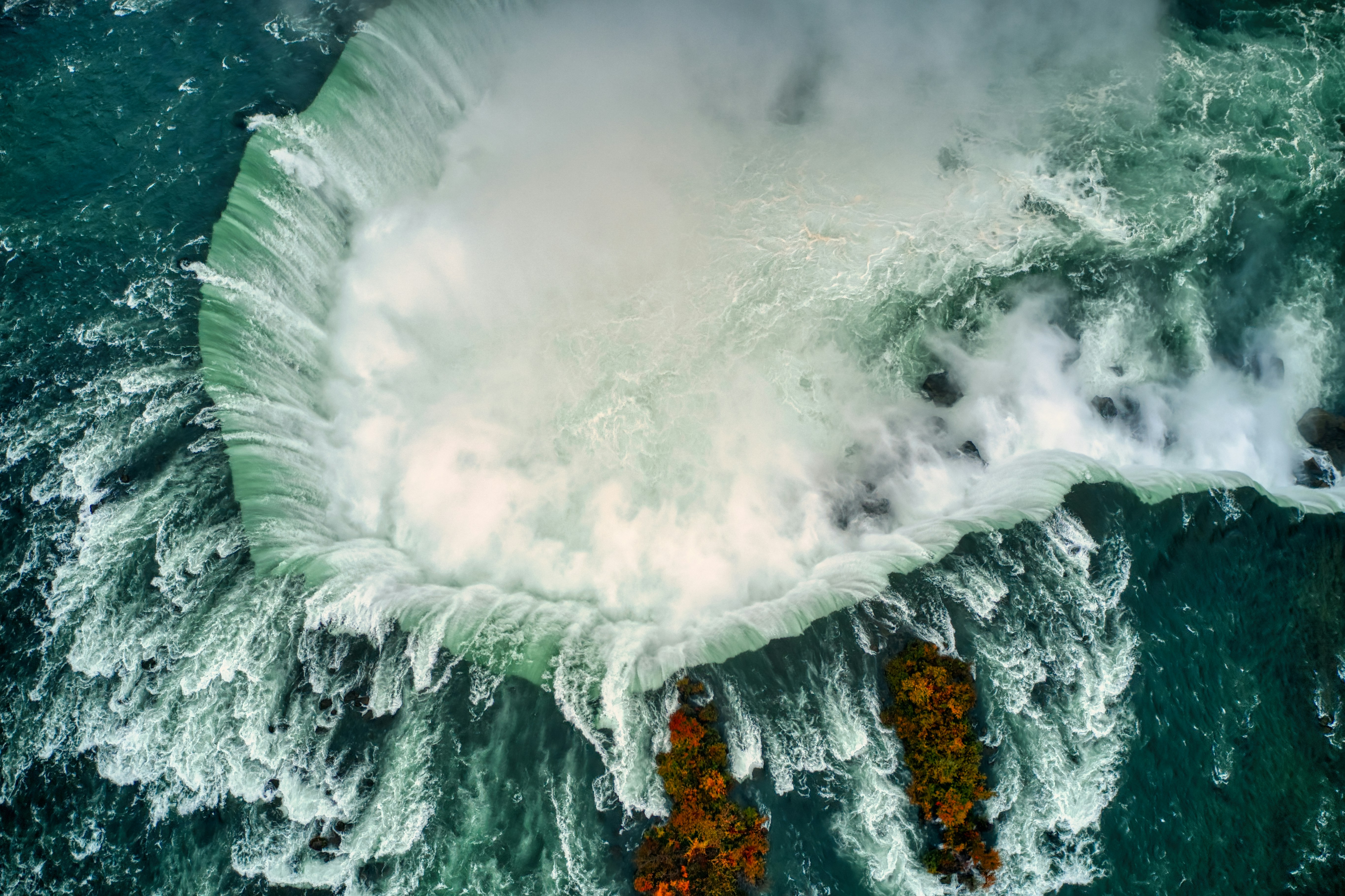 Aerial view of Niagara Falls' cascading waters with mist and vibrant autumn foliage.