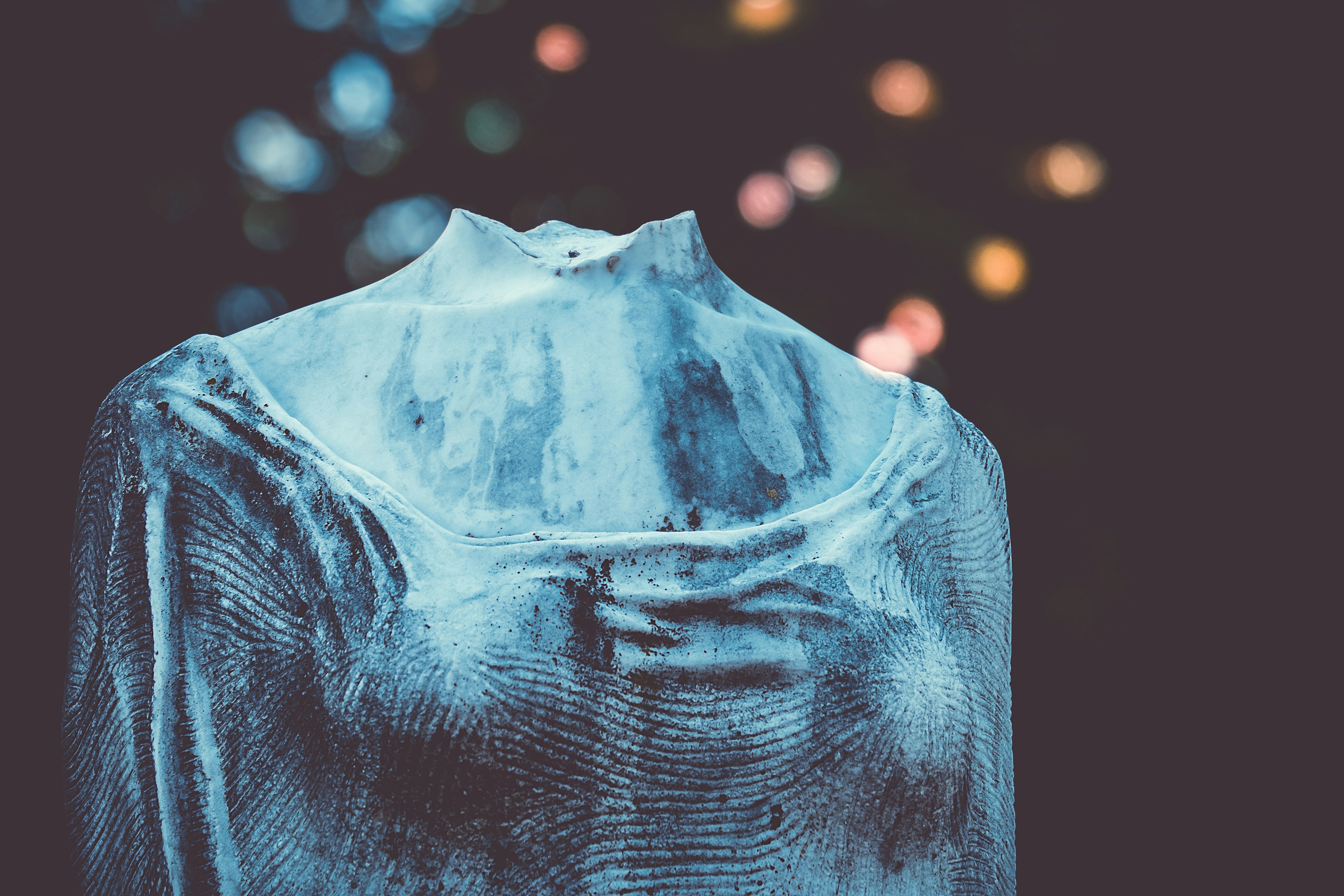 A sculptural torso devoid of a head, set against a softly blurred background of colorful lights. The texture and contours of the stone evoke a sense of timeless beauty.