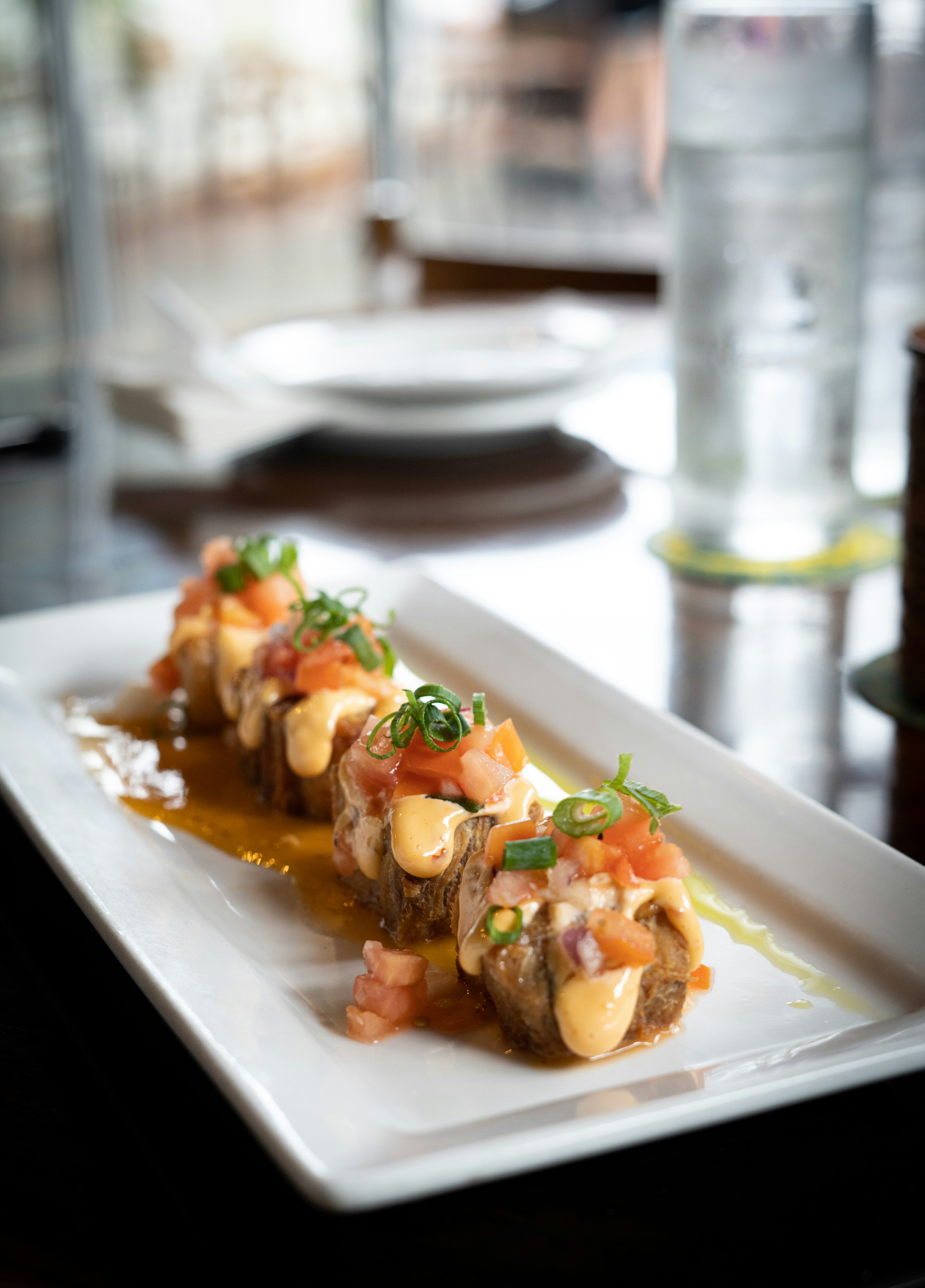 Delicious sushi rolls topped with fresh tomatoes and green onions, drizzled with a creamy sauce. The dish is elegantly presented on a white platter.