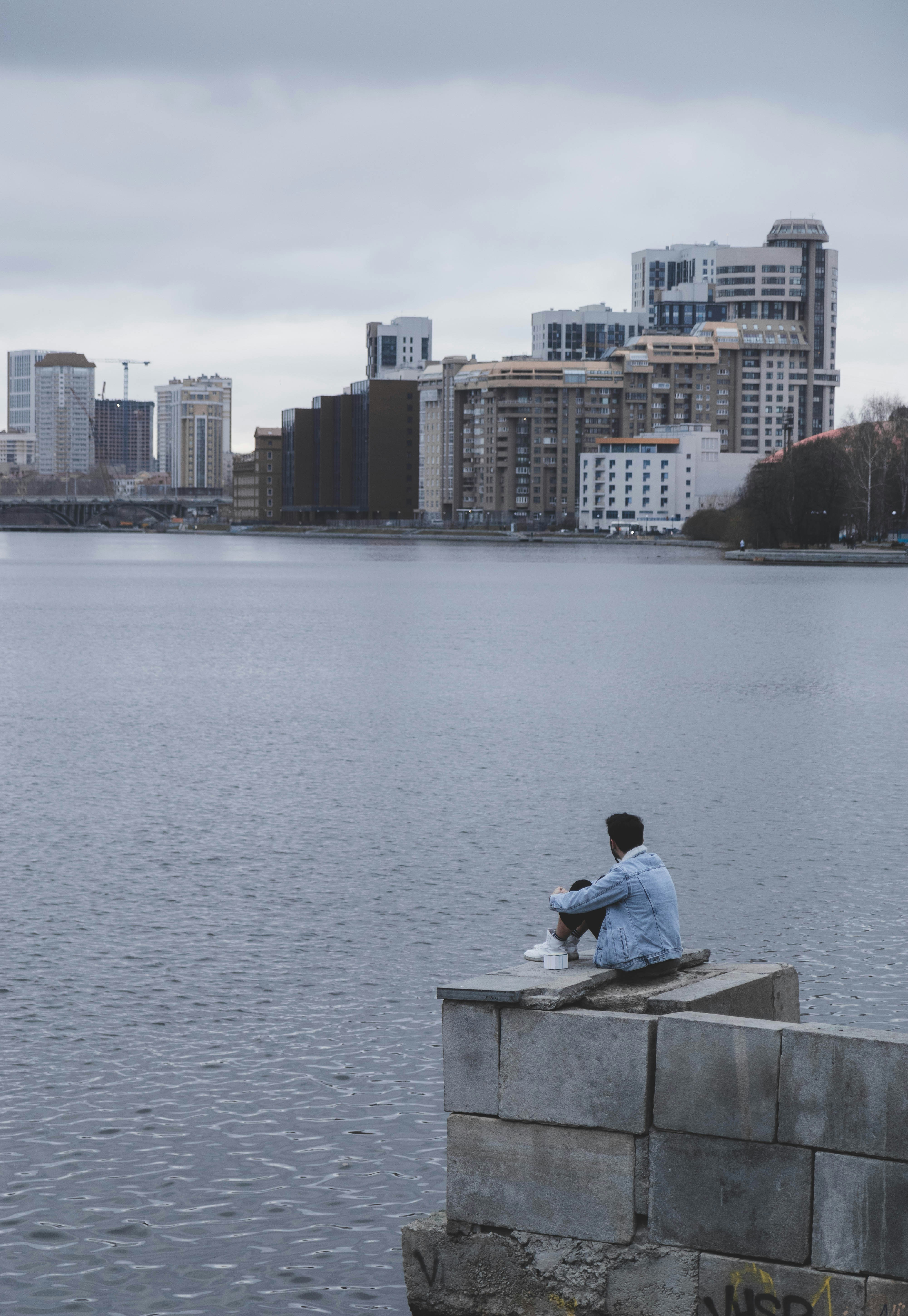 A lone figure sits on a stone ledge, gazing over a calm body of water with an urban skyline in the background. The scene captures a moment of reflection amidst the city.