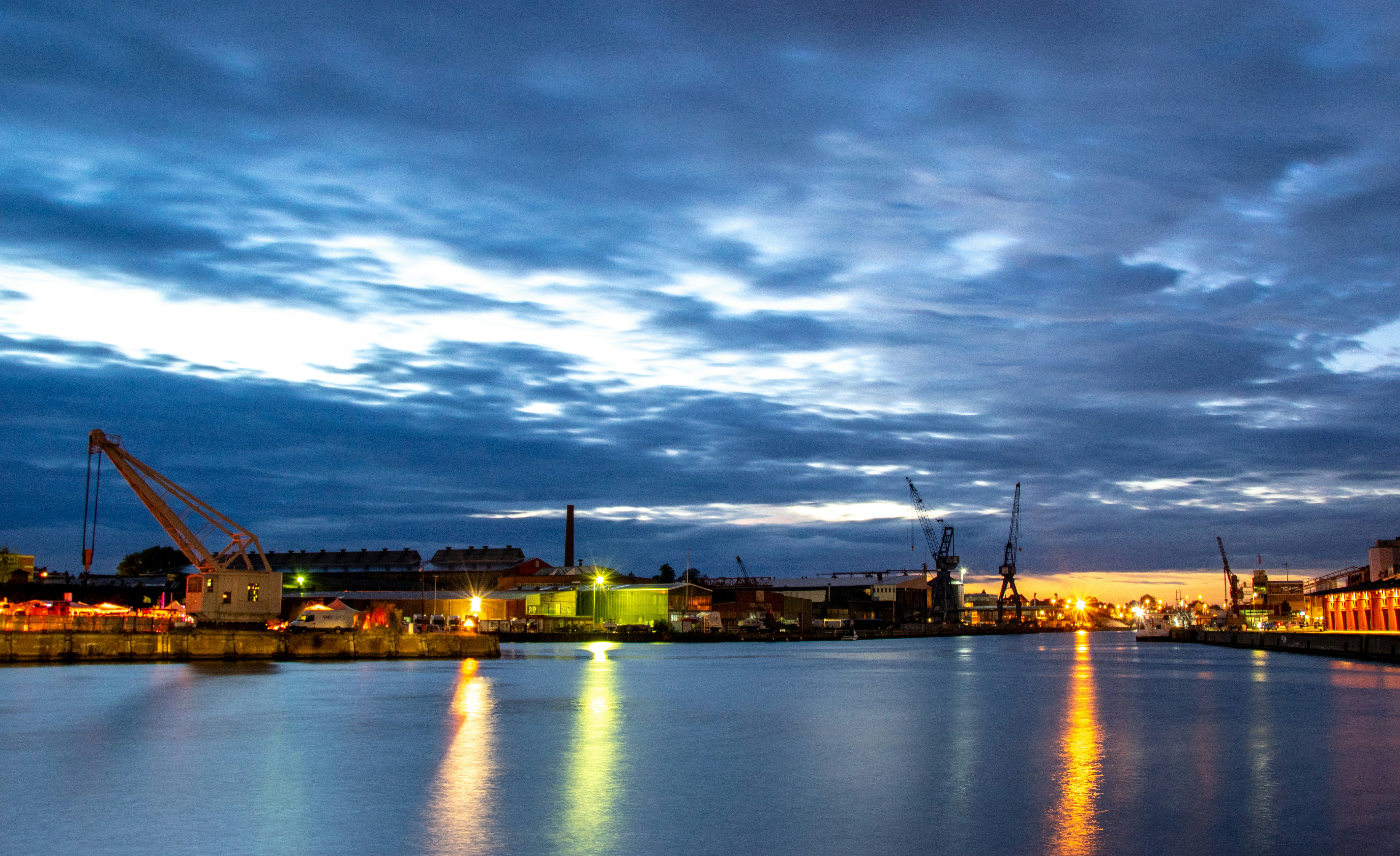 Harbor scene at dusk with illuminated cranes and buildings reflecting on calm water under a dramatic cloudy sky.