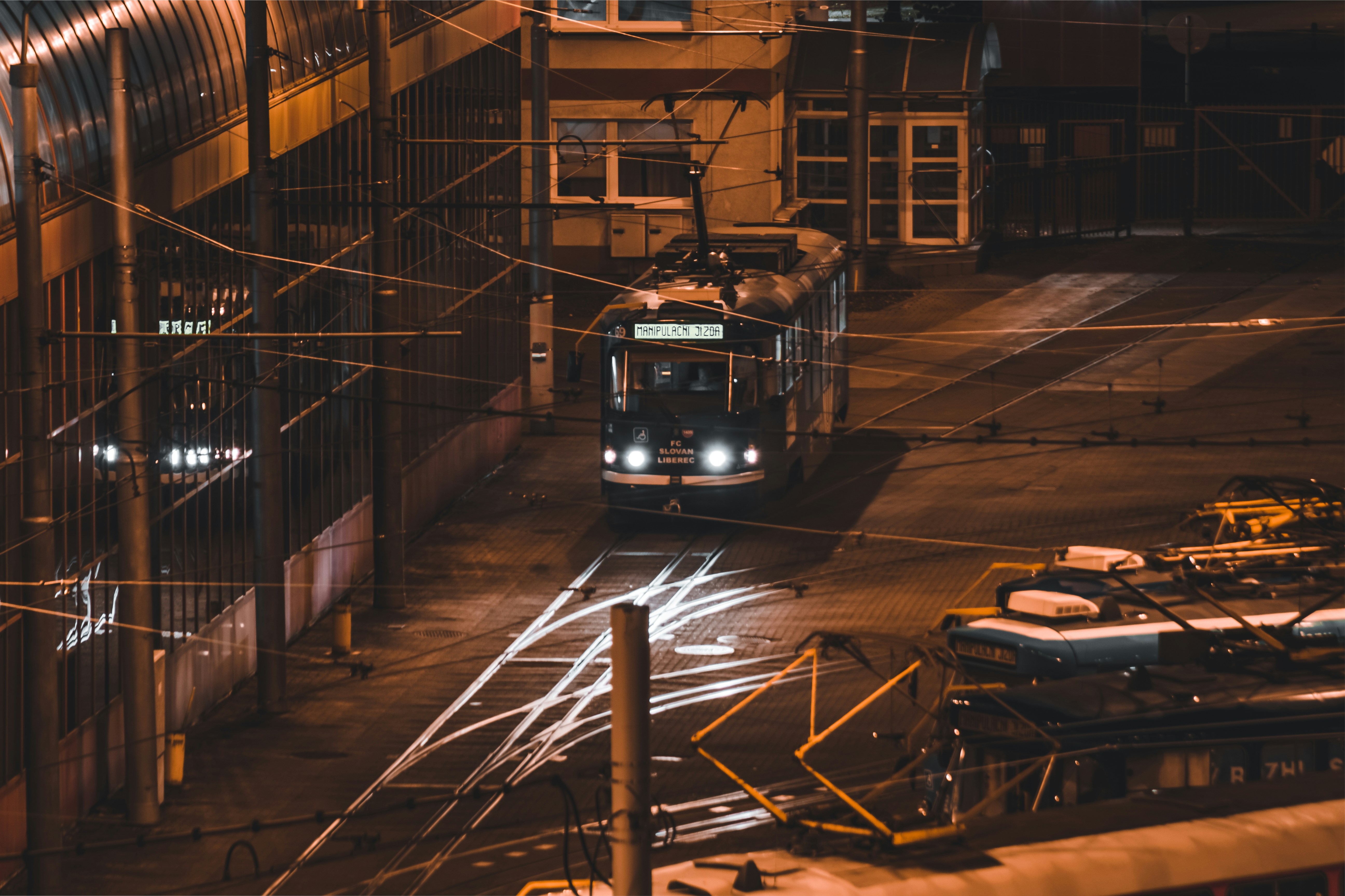 A tram navigates through a dimly lit depot, illuminated by scattered beams of light. The intricate network of tracks and buildings creates a dynamic urban scene.