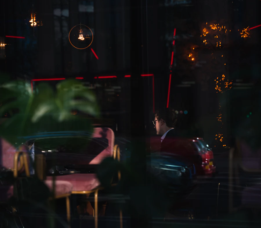 A dark, reflective café interior with warm hanging lights, red neon lines, and a man seated in profile near the right side. Blurred greenery and chairs add depth in the foreground.
