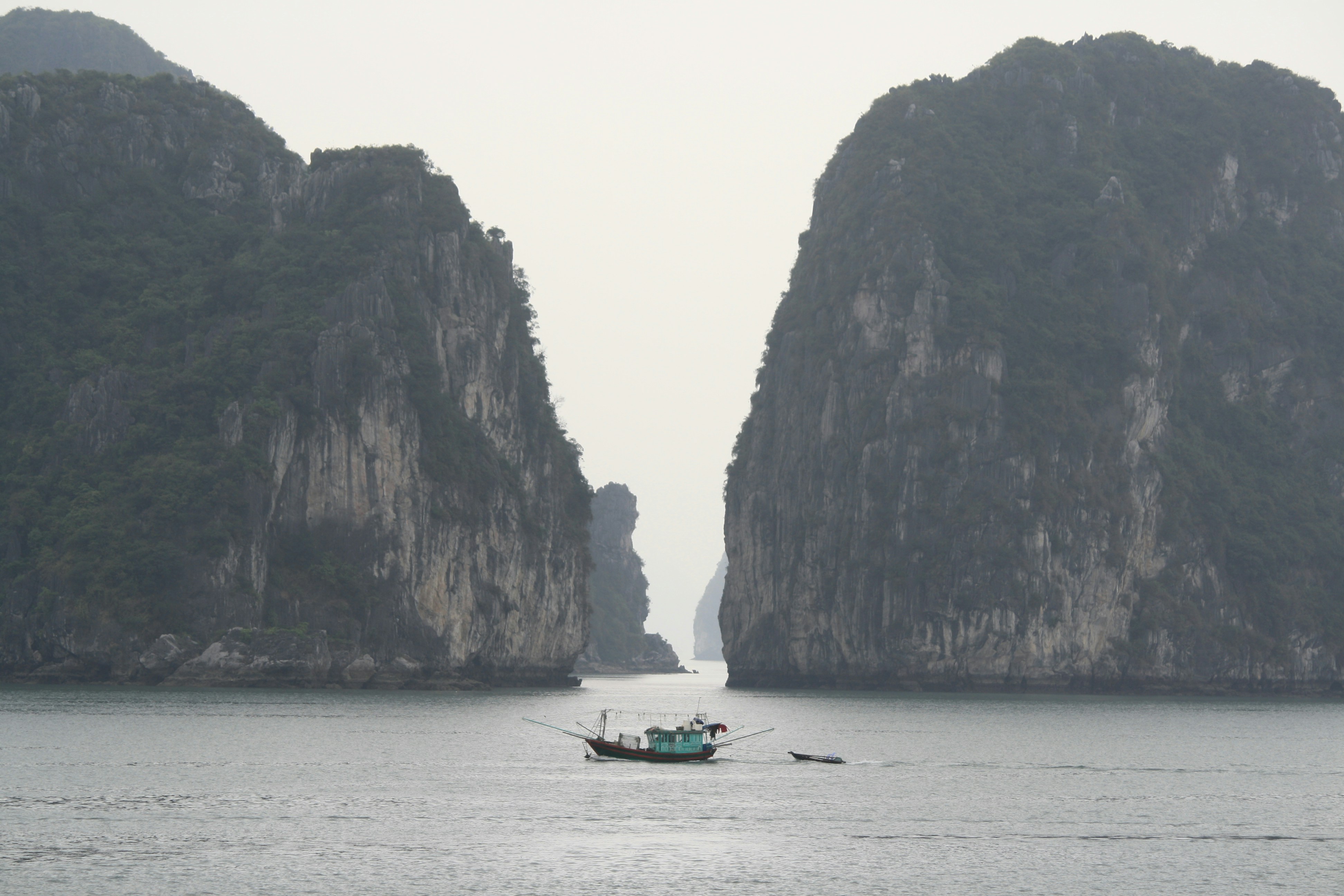 Traditional boat navigating through a narrow passage between towering limestone cliffs in a misty seascape.