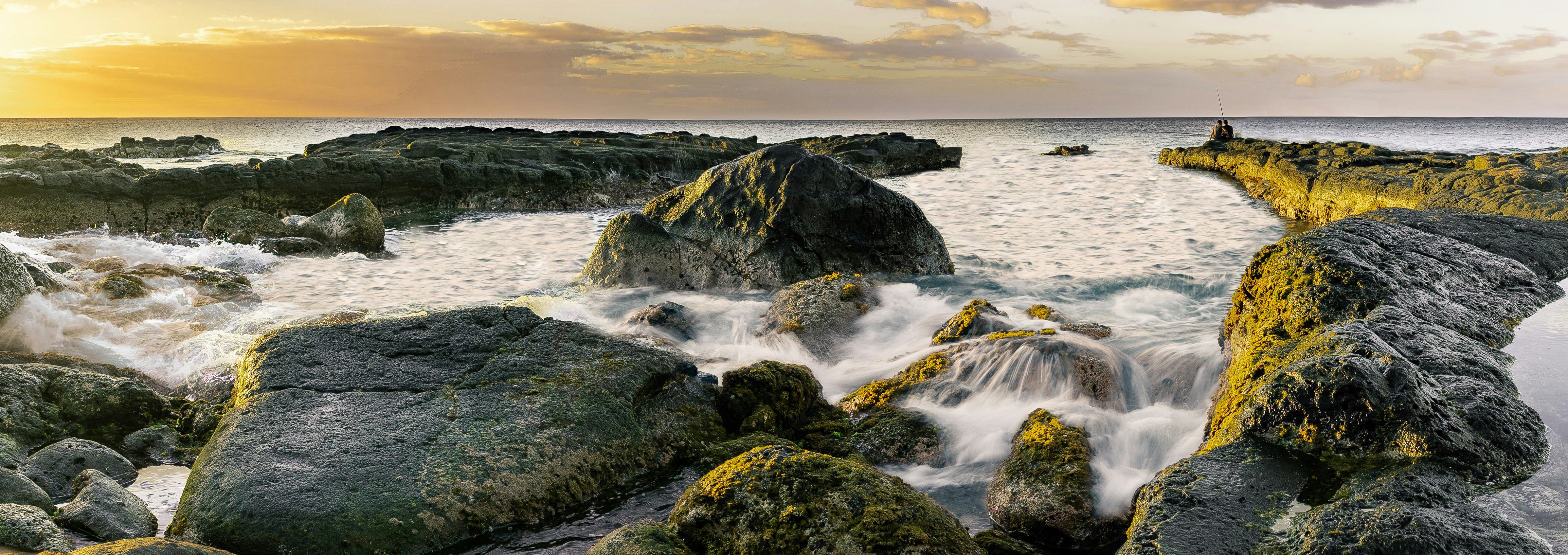 Waves gently crashing over moss-covered rocks at sunset near a distant lighthouse.