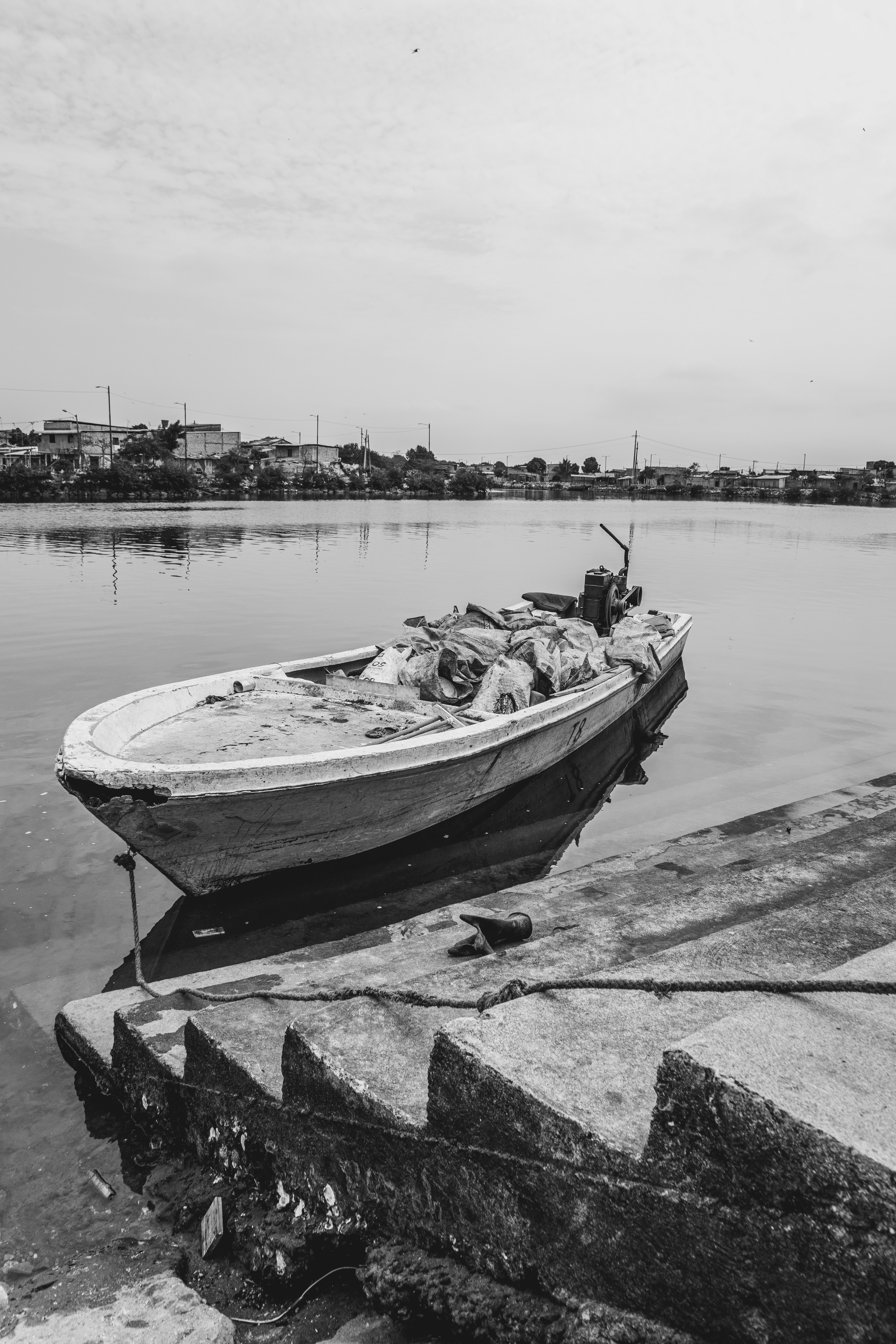 Grayscale view of a weathered boat moored at a calm waterfront with distant urban skyline.