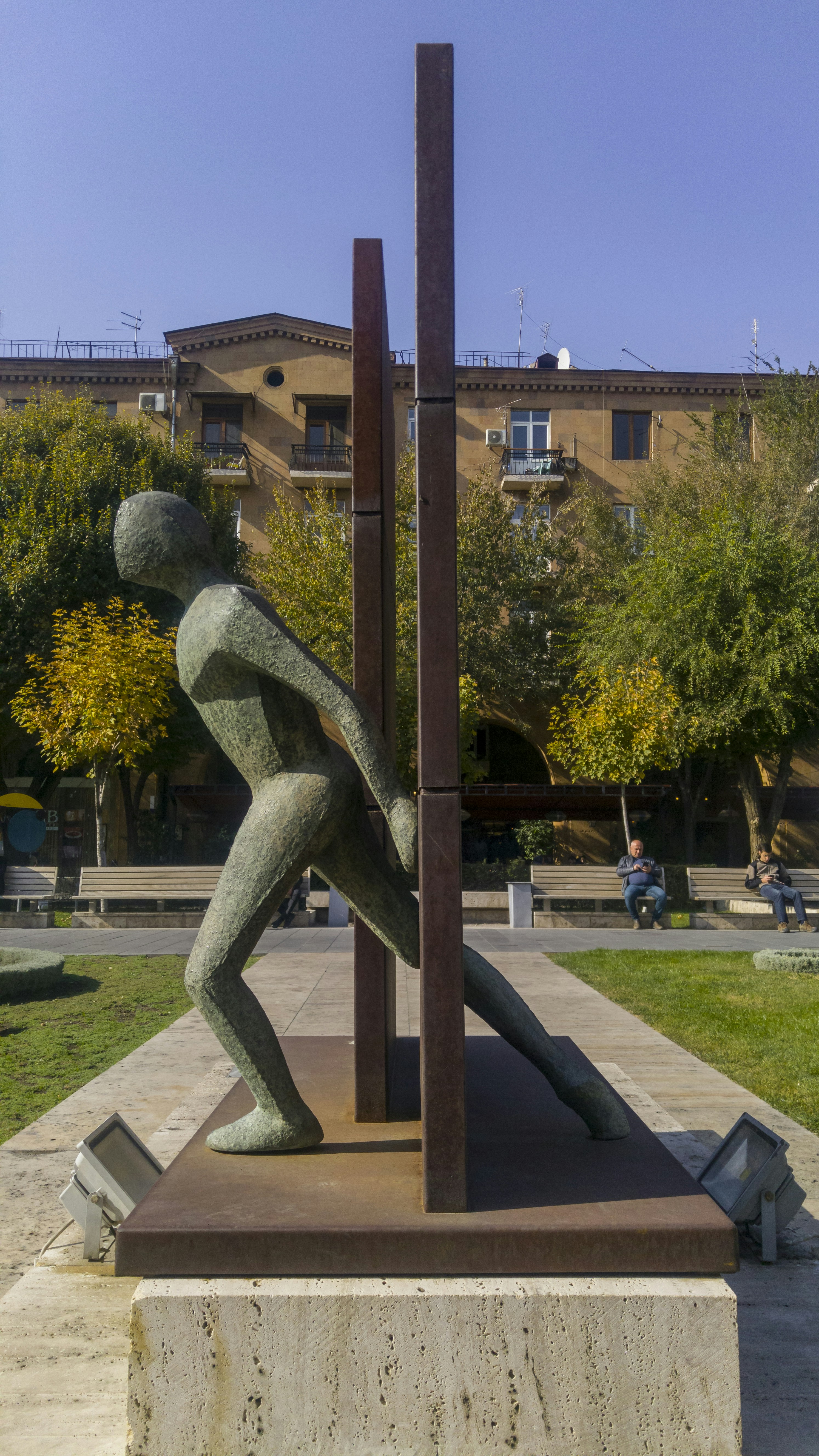 A dynamic sculpture of a figure straining against vertical bars, set in a vibrant urban park with autumn foliage. The scene captures a moment of tension and resilience.
