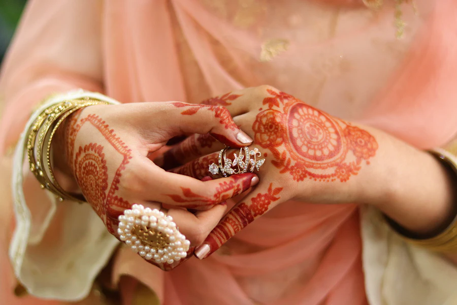 Close-up of a woman’s hands decorated with intricate henna, holding a jeweled ring. Soft peach clothing and gold bangles frame the ornate details.