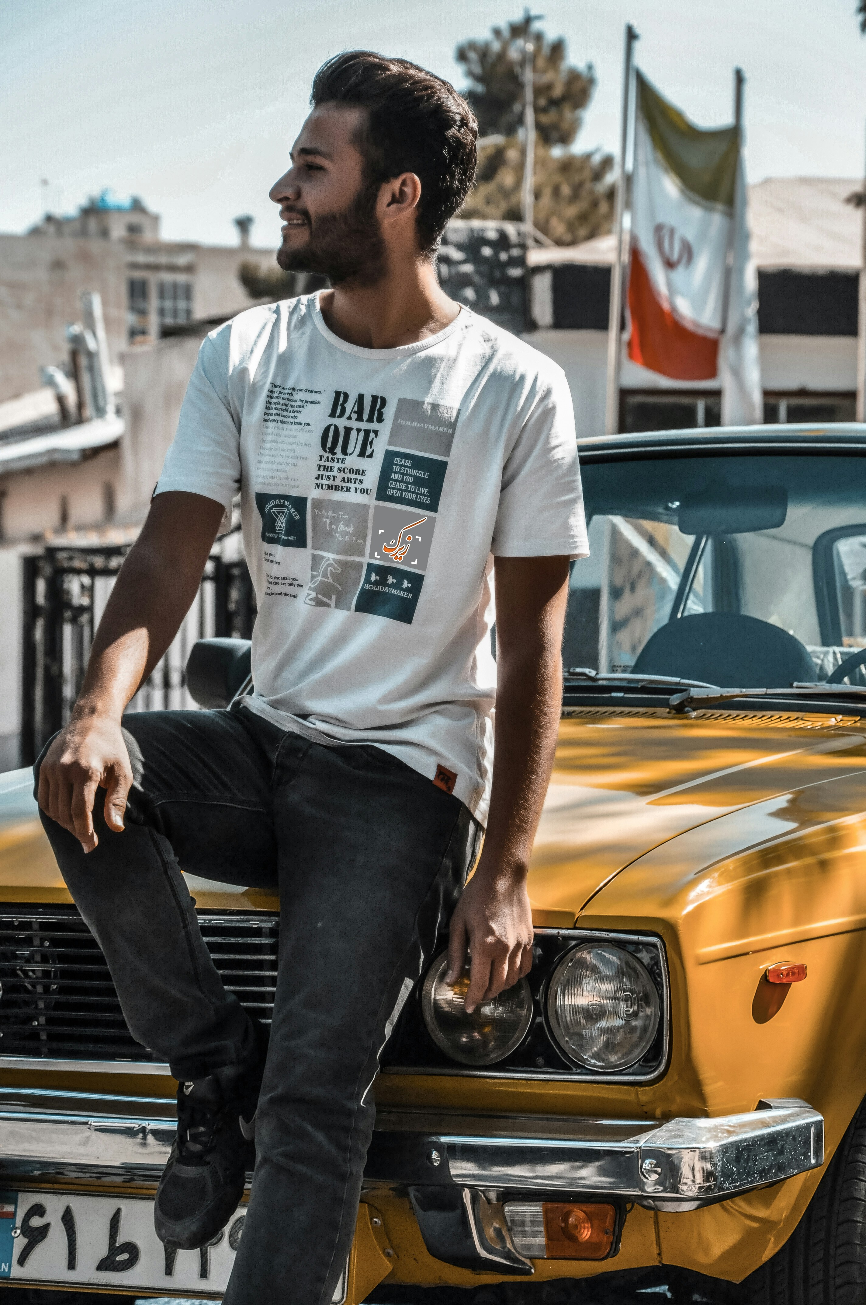 Young man casually leaning on a vintage yellow car, showcasing a playful graphic tee against a backdrop of flags and urban architecture.